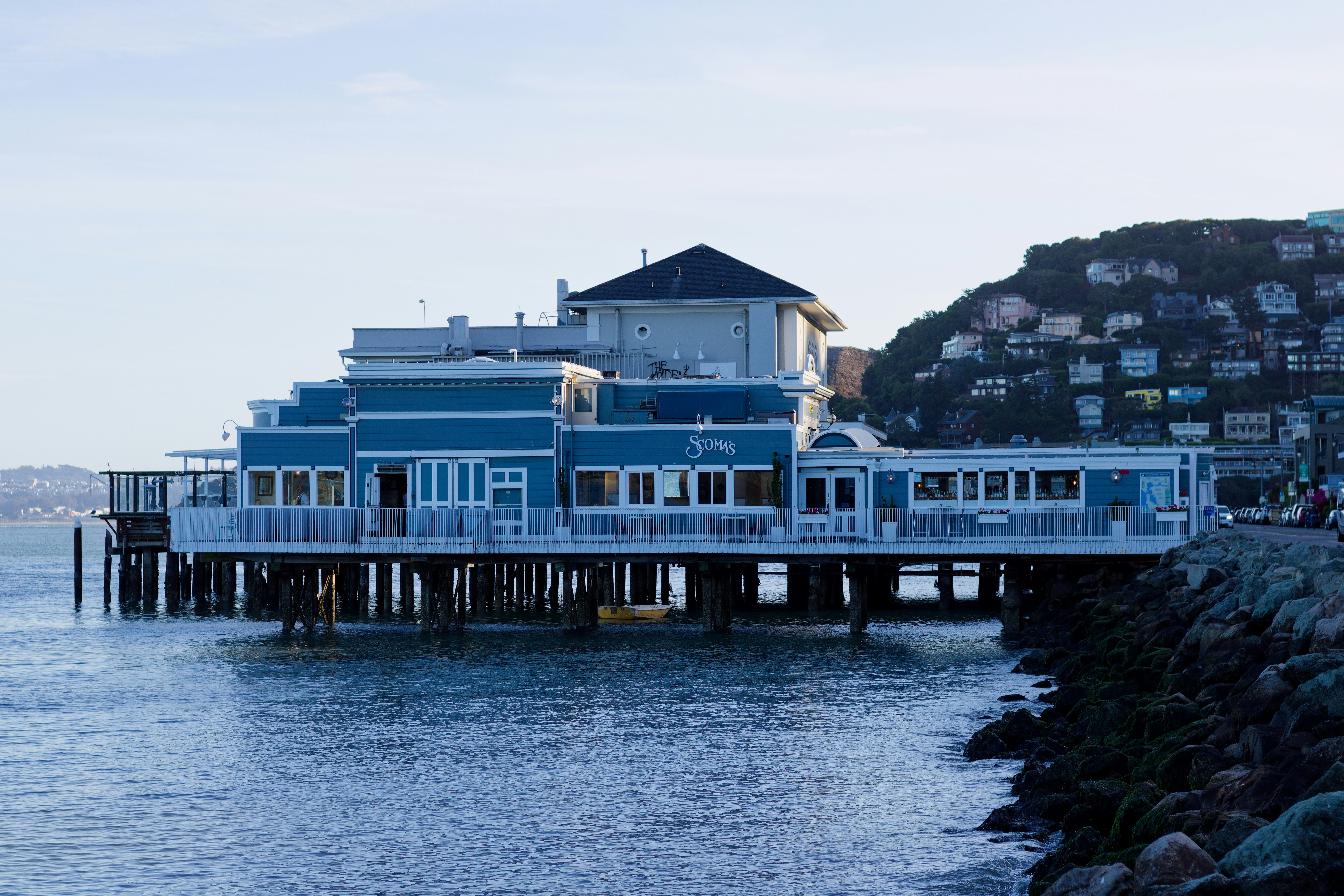 Blue-toned waterfront restaurant on a pier extending over calm sea, with rocky jetty on the right and hillside housing in the background.