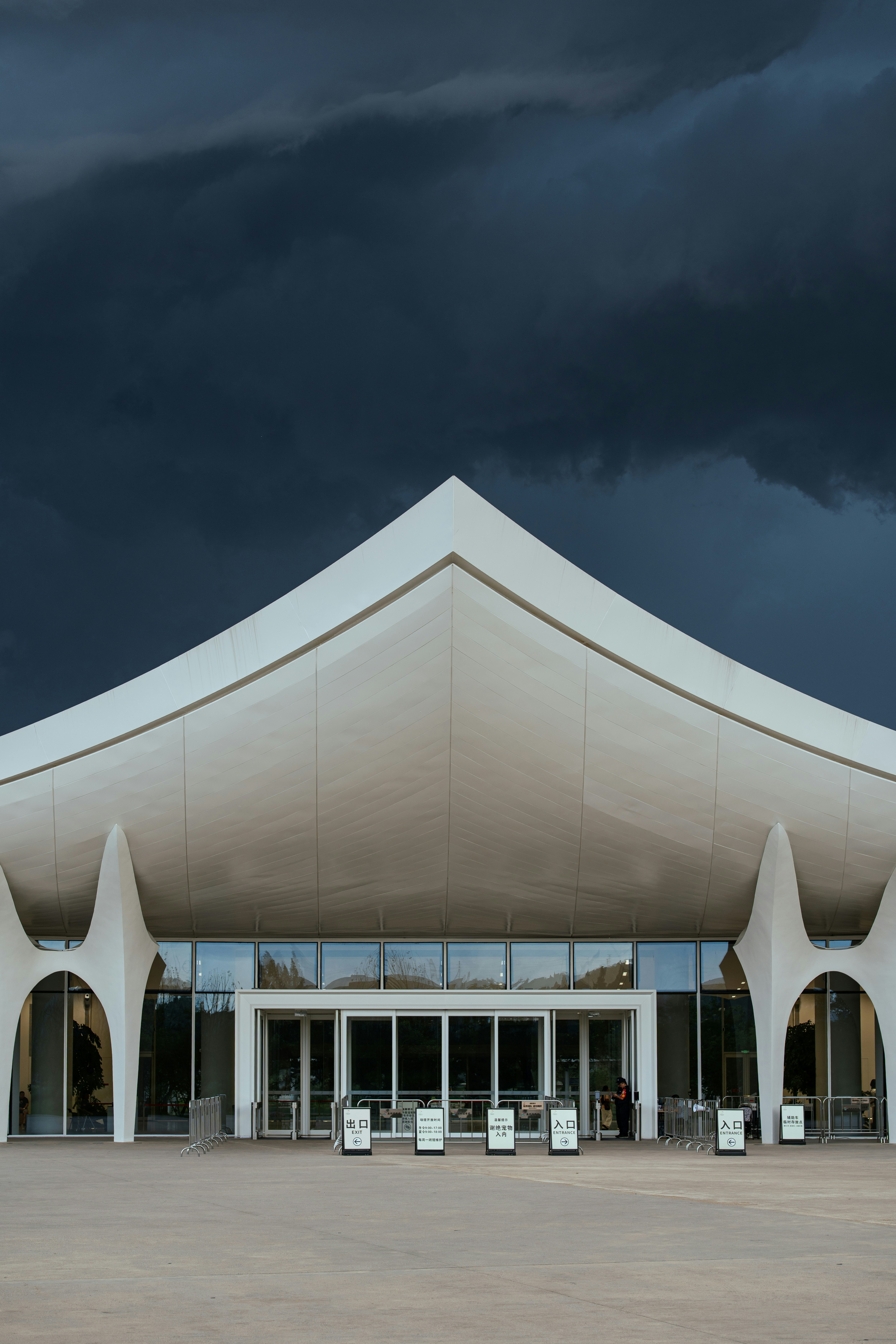 A modern building entrance with a sweeping curved roof beneath a dramatic dark sky; the glass facade and open plaza convey a ceremonial, institutional vibe.