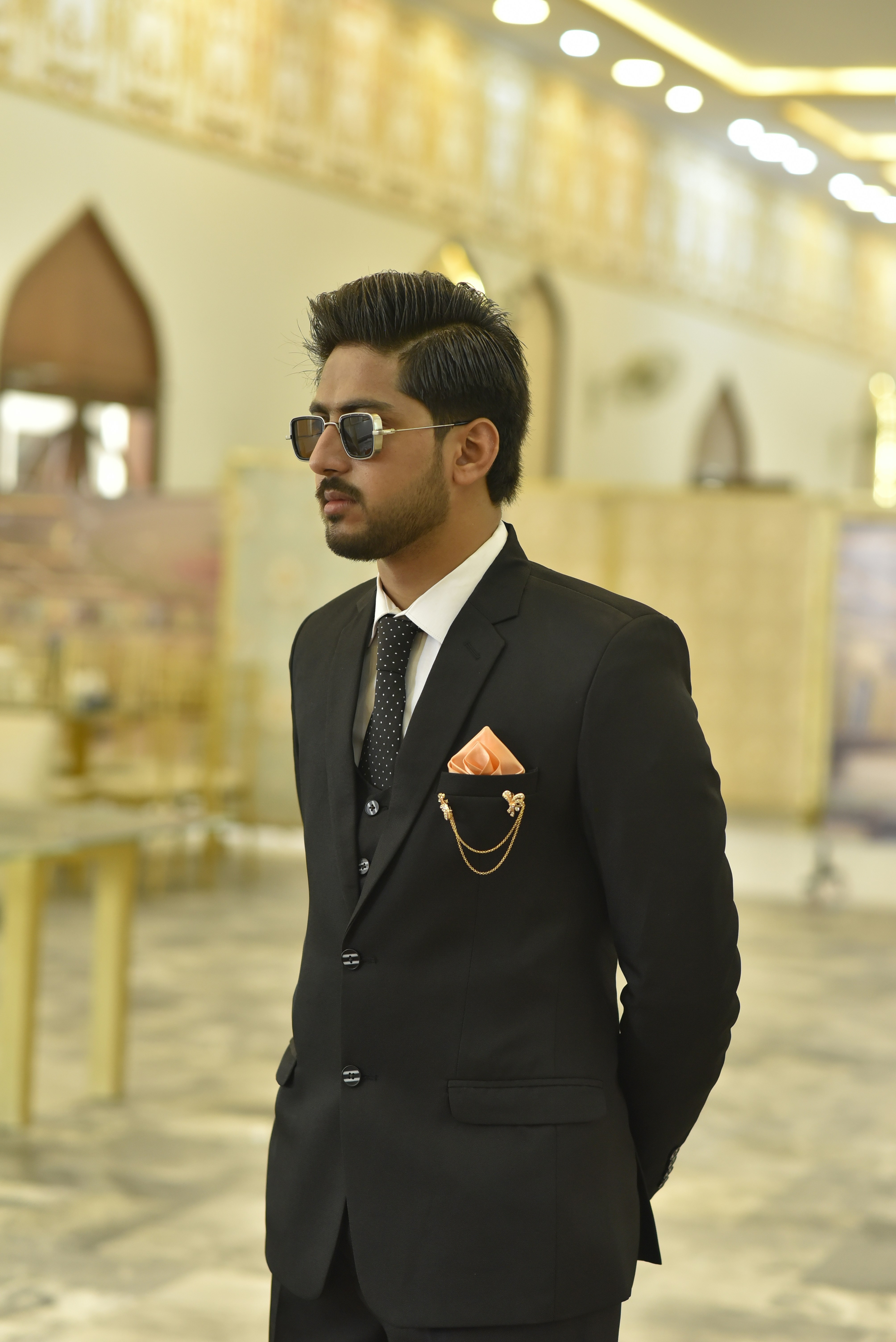 Dapper silhouette: a sharply dressed man in a tailored black suit stands poised in an ornate, softly lit hall