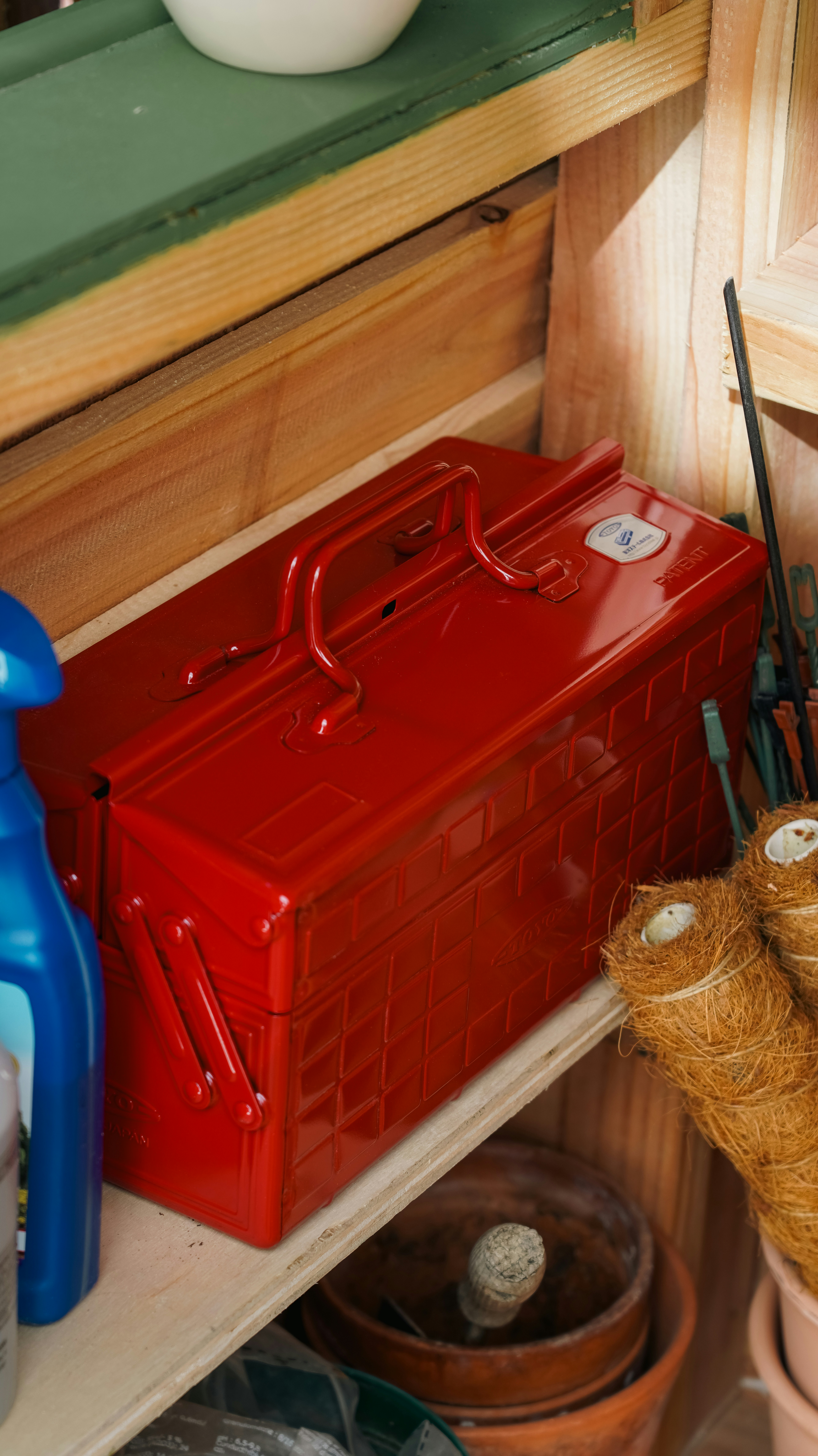 Red toolbox on a wooden shelf in a cluttered shed, with tools and rope visible nearby.