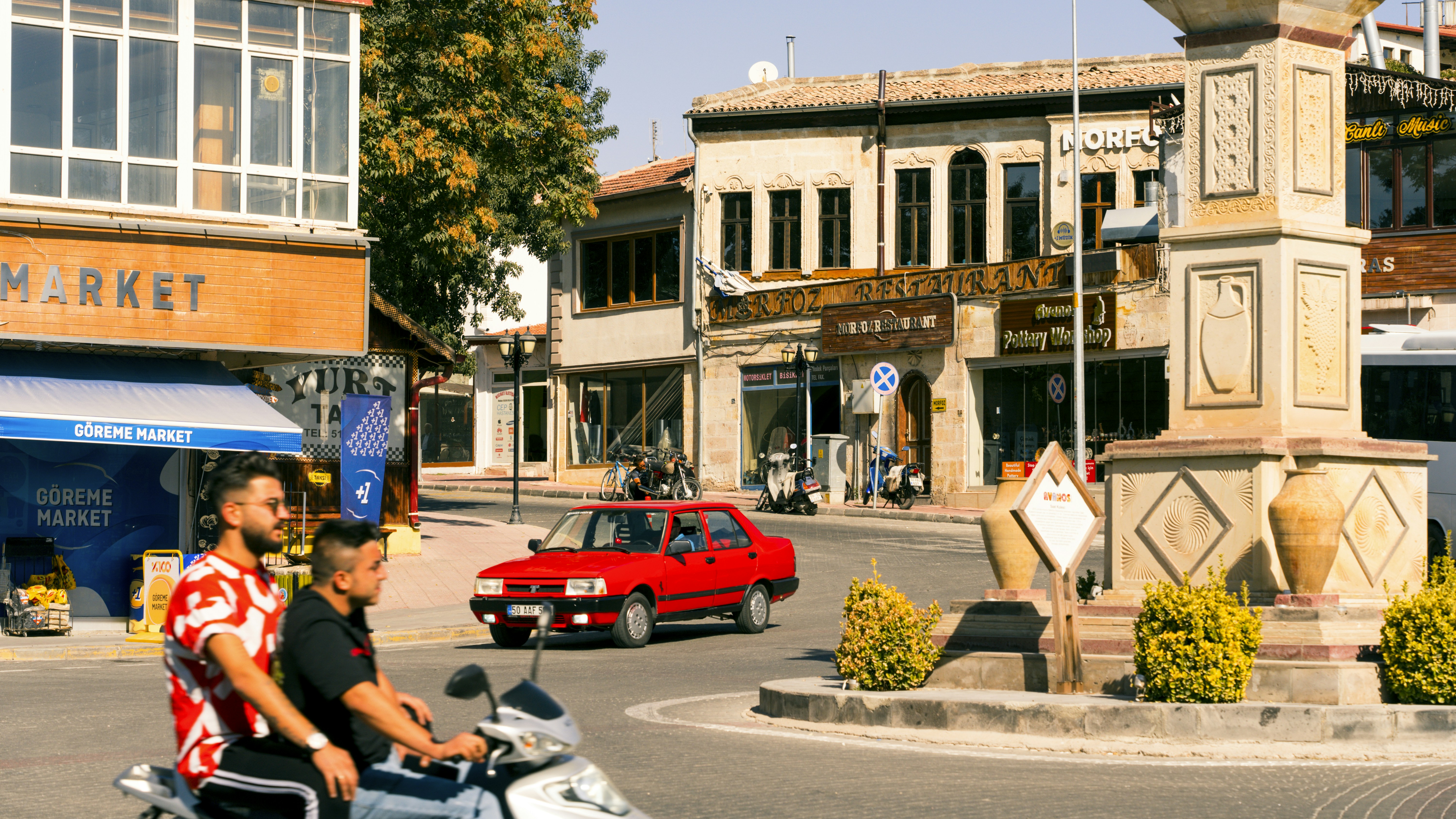 Street Roundabout Chorus: A Red Car, Stone Monument, and Market Front in Afternoon Light