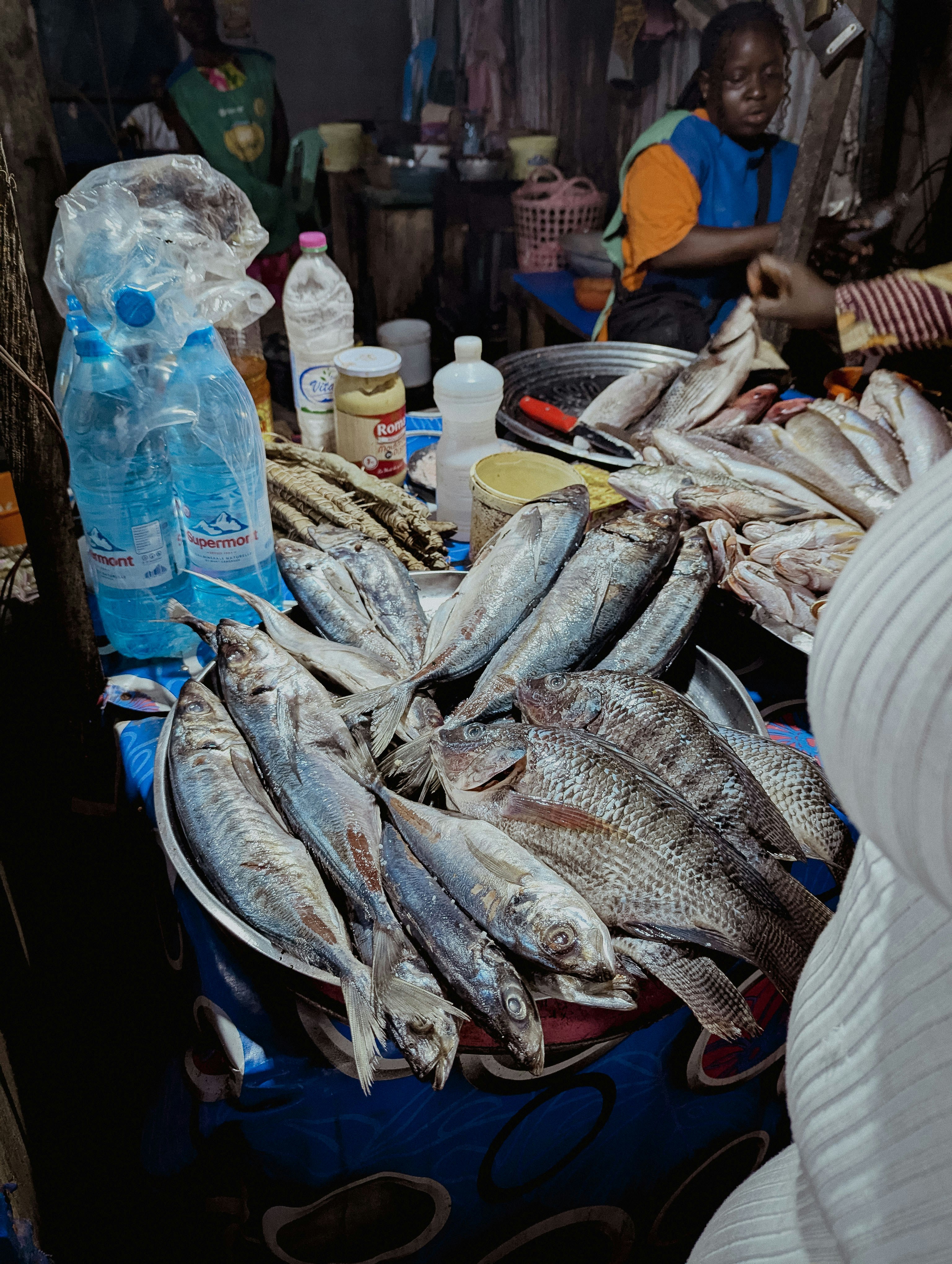 Fresh fish piled on ice at a busy market stall, with a vendor in the background and assorted bottles behind. A candid street-food scene showing texture and color variety; not a studio shot, with natural lighting.
