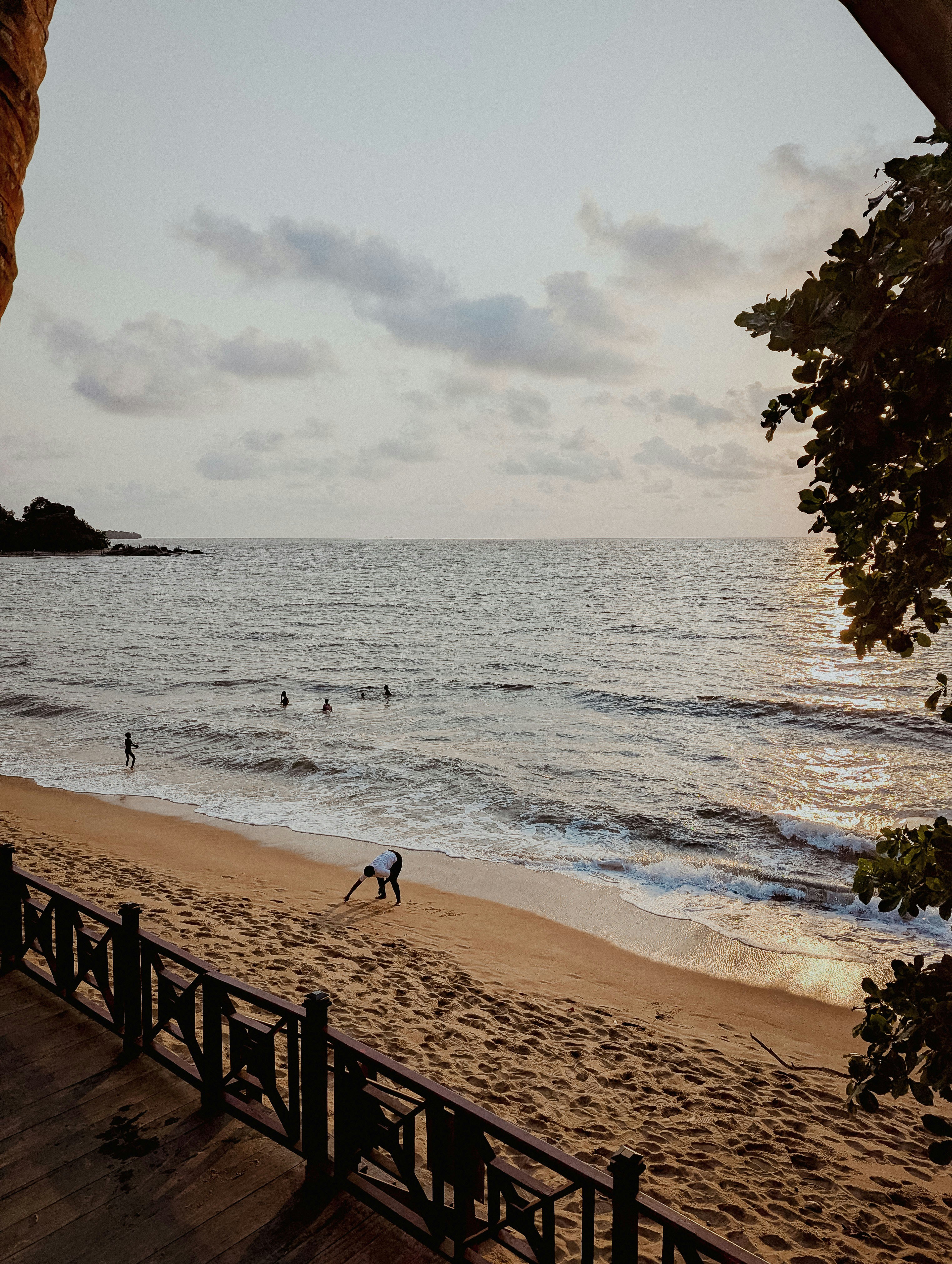 Golden beach at dusk with a lone person digging in the sand while a calm sea stretches to the horizon; silhouetted figures in the water and a wooden railing framing the foreground.