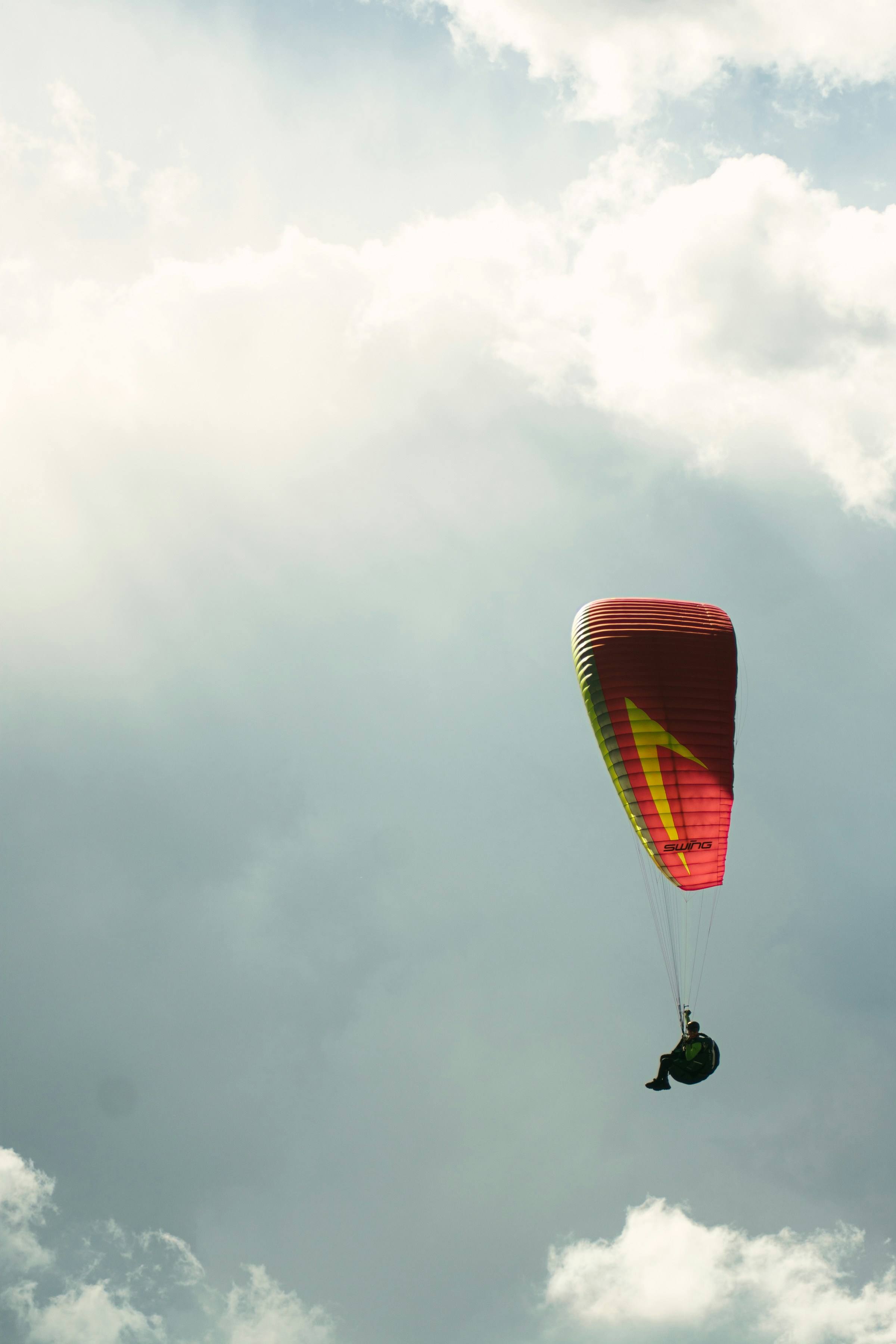 Red paraglider sails through pale clouds as a lone pilot drifts in daylight