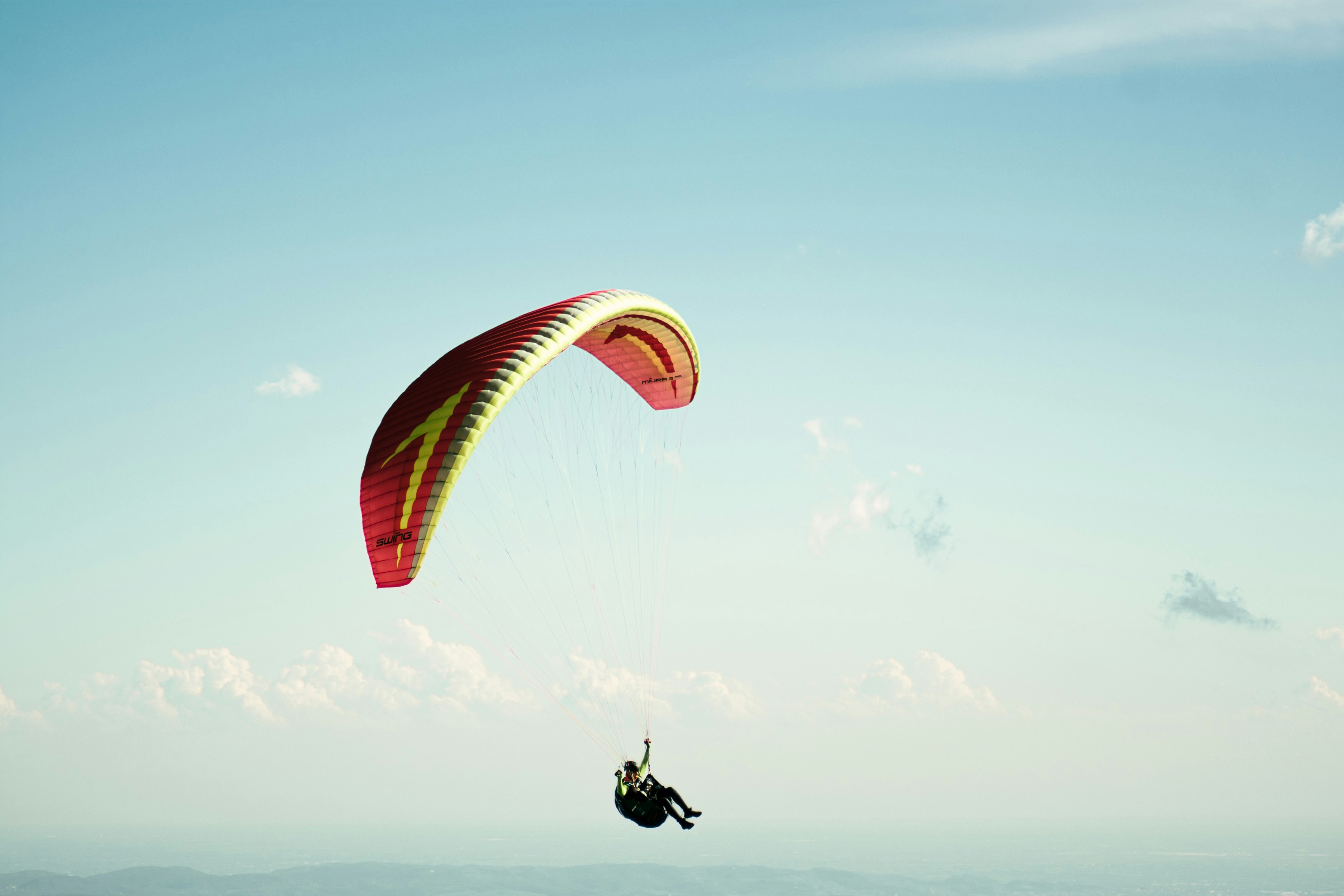 A lone paraglider rides a red and yellow kite over a calm horizon, set against a pale blue sky with soft clouds. The scene emphasizes flight and freedom without clutter or crowd.