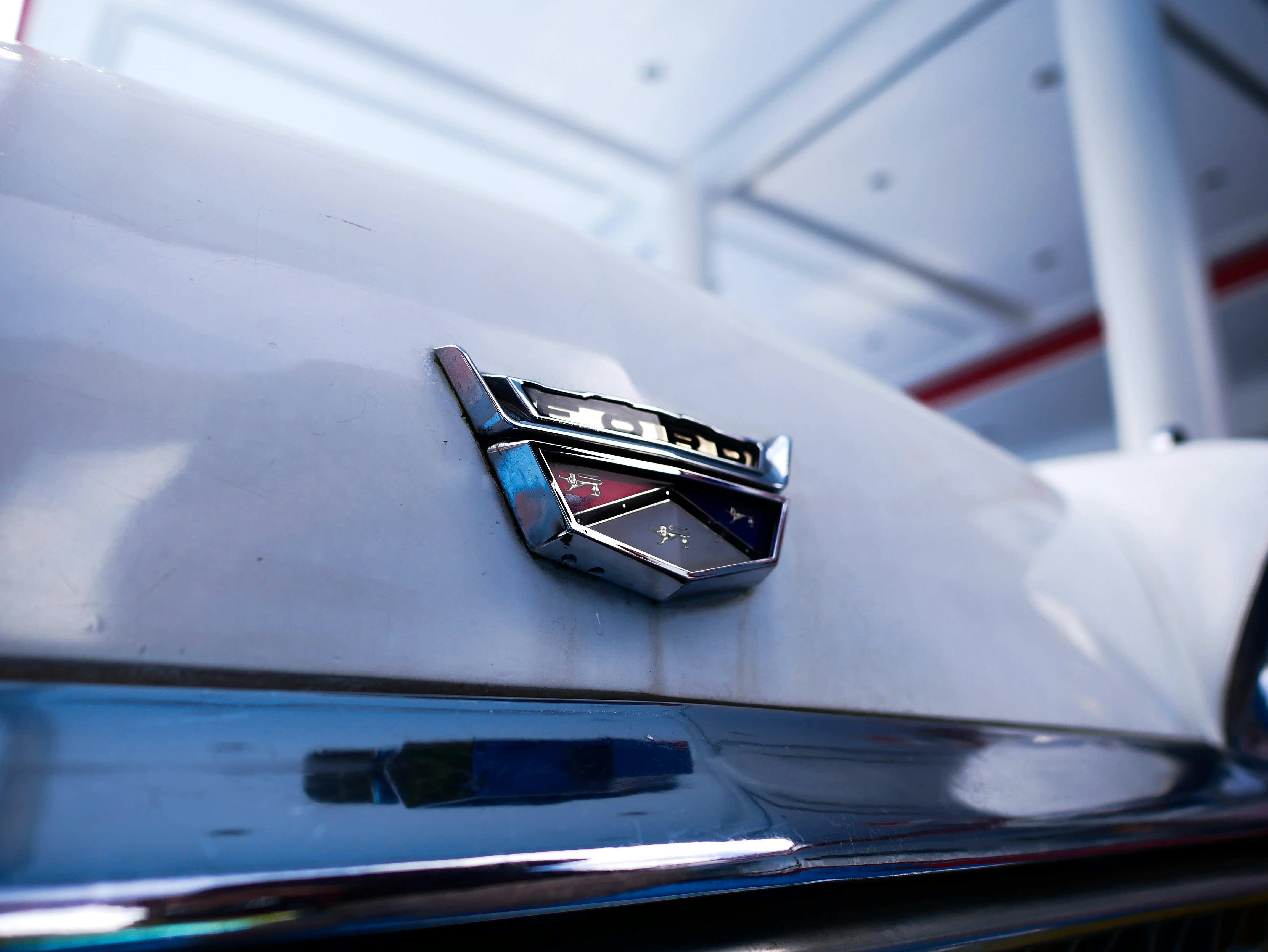 Close-up of a vintage car badge on a pale body with blue chrome trim, under a white, industrial canopy; emphasizes logo and surface reflections.