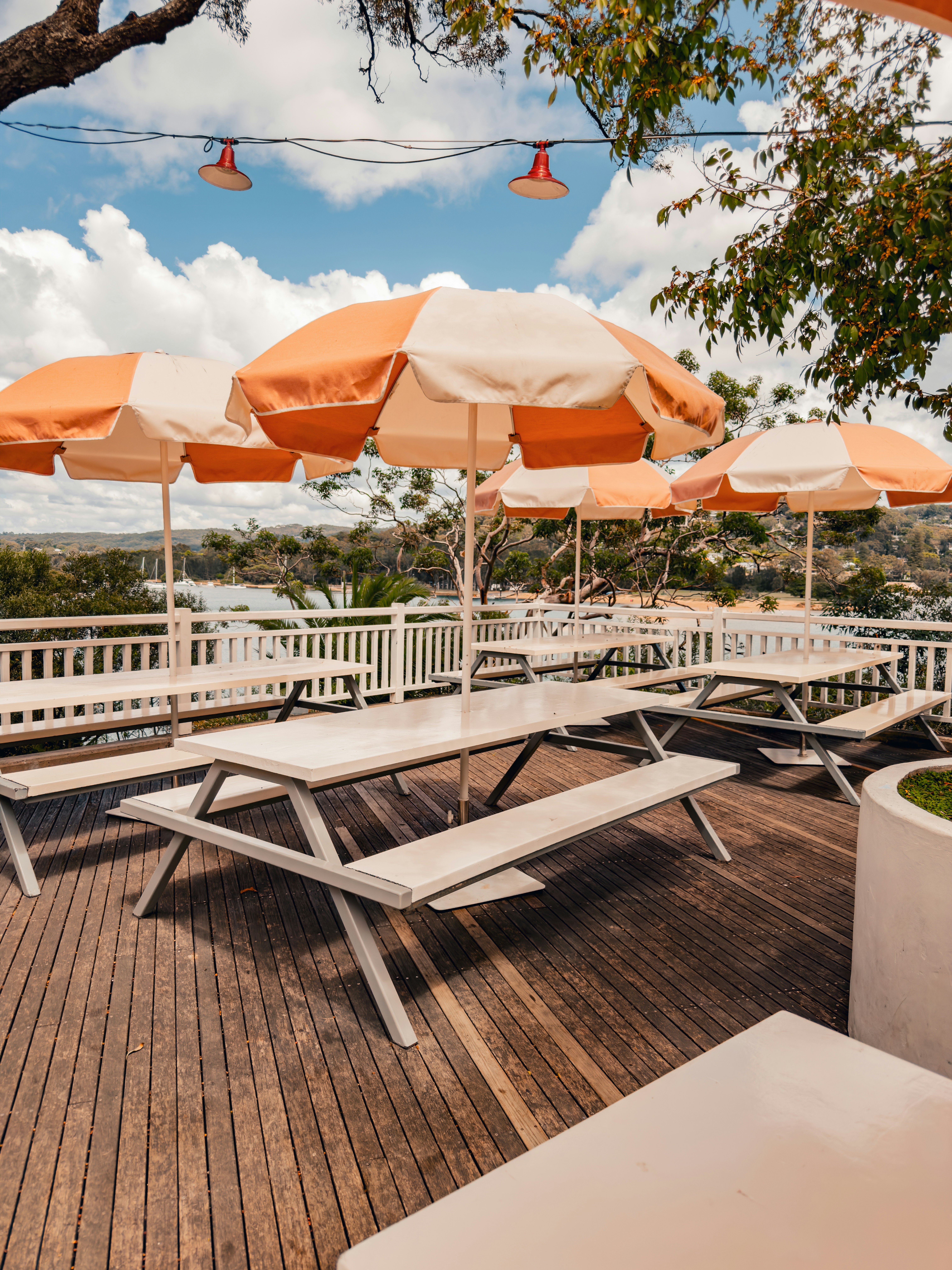 Sunny rooftop deck with orange and cream umbrellas shading wooden picnic tables and benches; distant landscape and blue sky with clouds in the background.