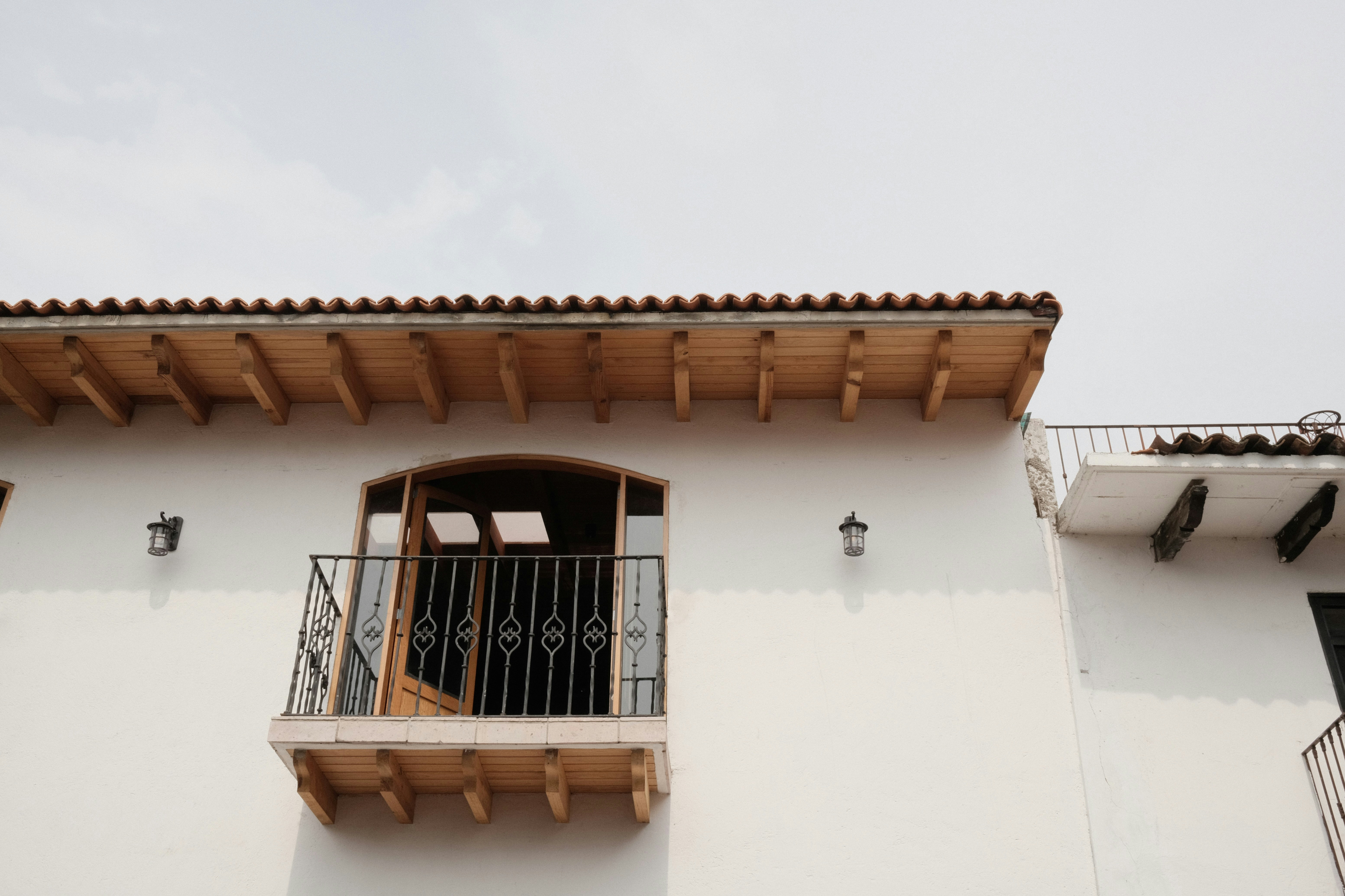 White stucco building façade with a wooden balcony and iron railing, set under a terracotta tiled roof; minimal sky visible.