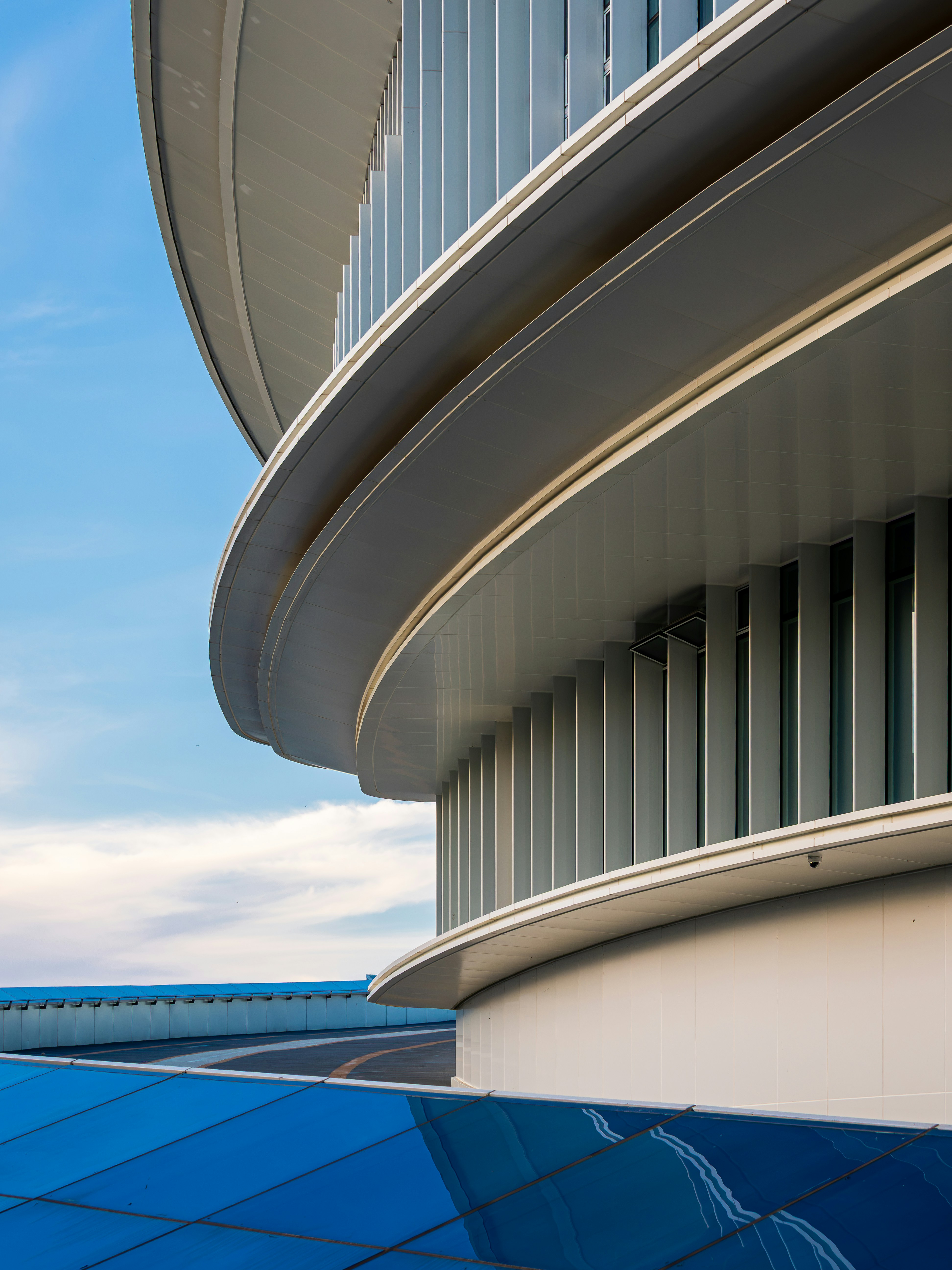Curved architectural concrete and glass forms stack in sweeping, concentric lines against a blue sky, with reflective blue panels at the base adding a bold color contrast.