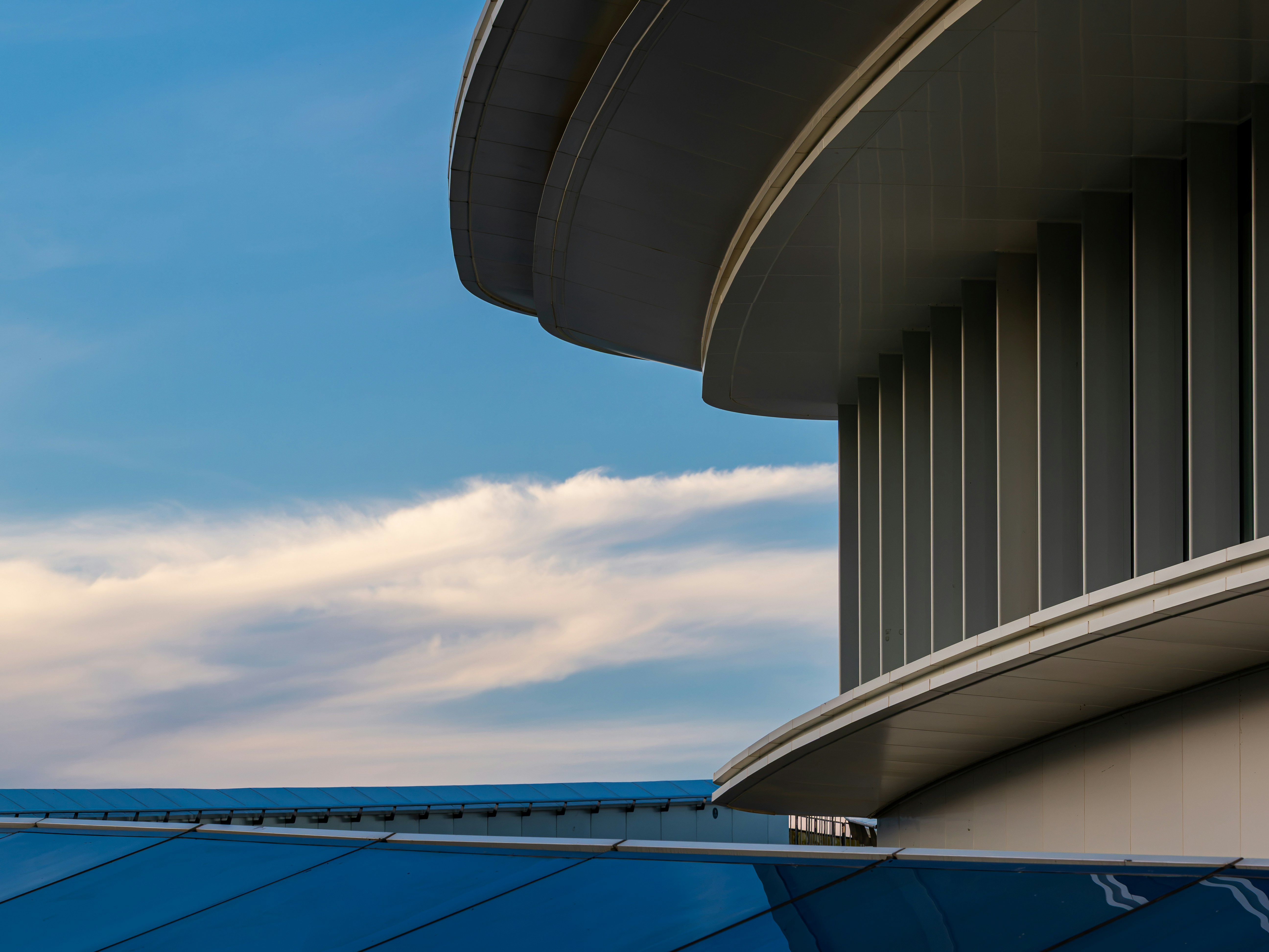 Architectural curve forms and rounded eaves frame a modern building against a blue sky with wispy clouds. The image emphasizes the contrast between sweeping architectural geometry and clean lines of the facade.