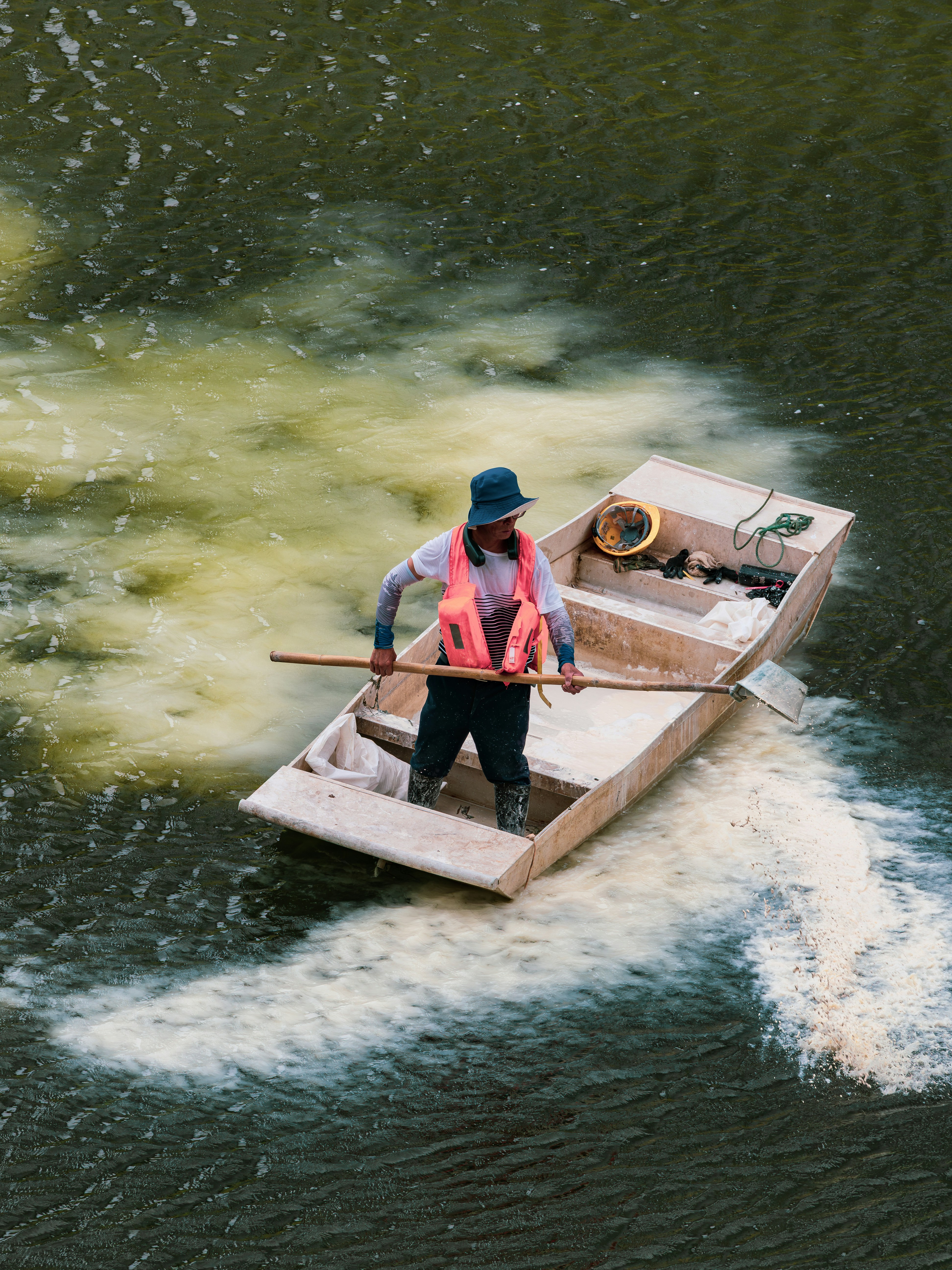 A fisherman stands in a small wooden boat navigating churned river water, wearing a blue hat and life jacket as he rows with a long paddle; the scene emphasizes movement and resilience in a narrow craft.