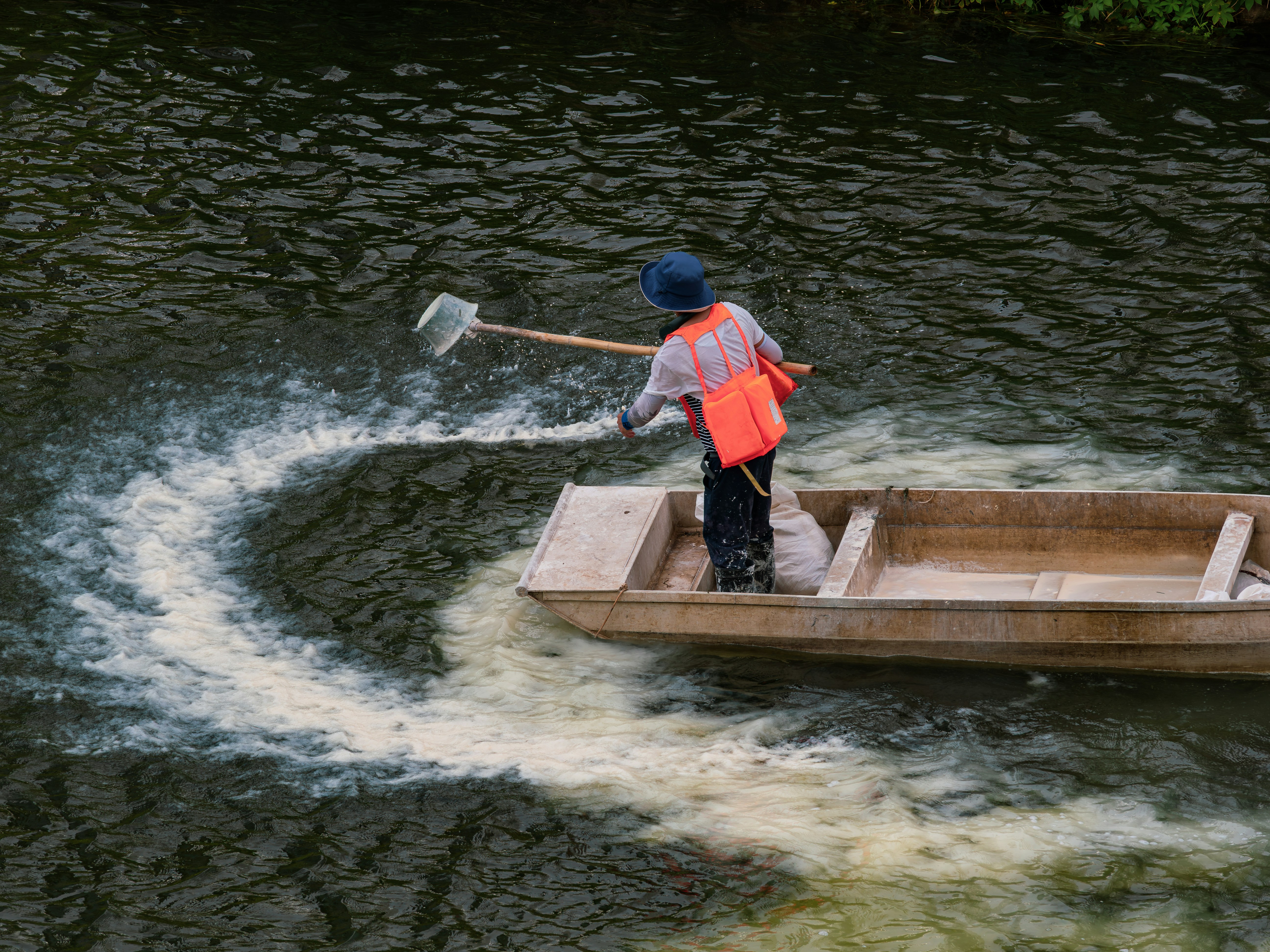 A person in a wooden boat casts a sharp splash of water with a long-handled paddle in a river setting; bright life jacket and dark hat stand out against the rippling water.