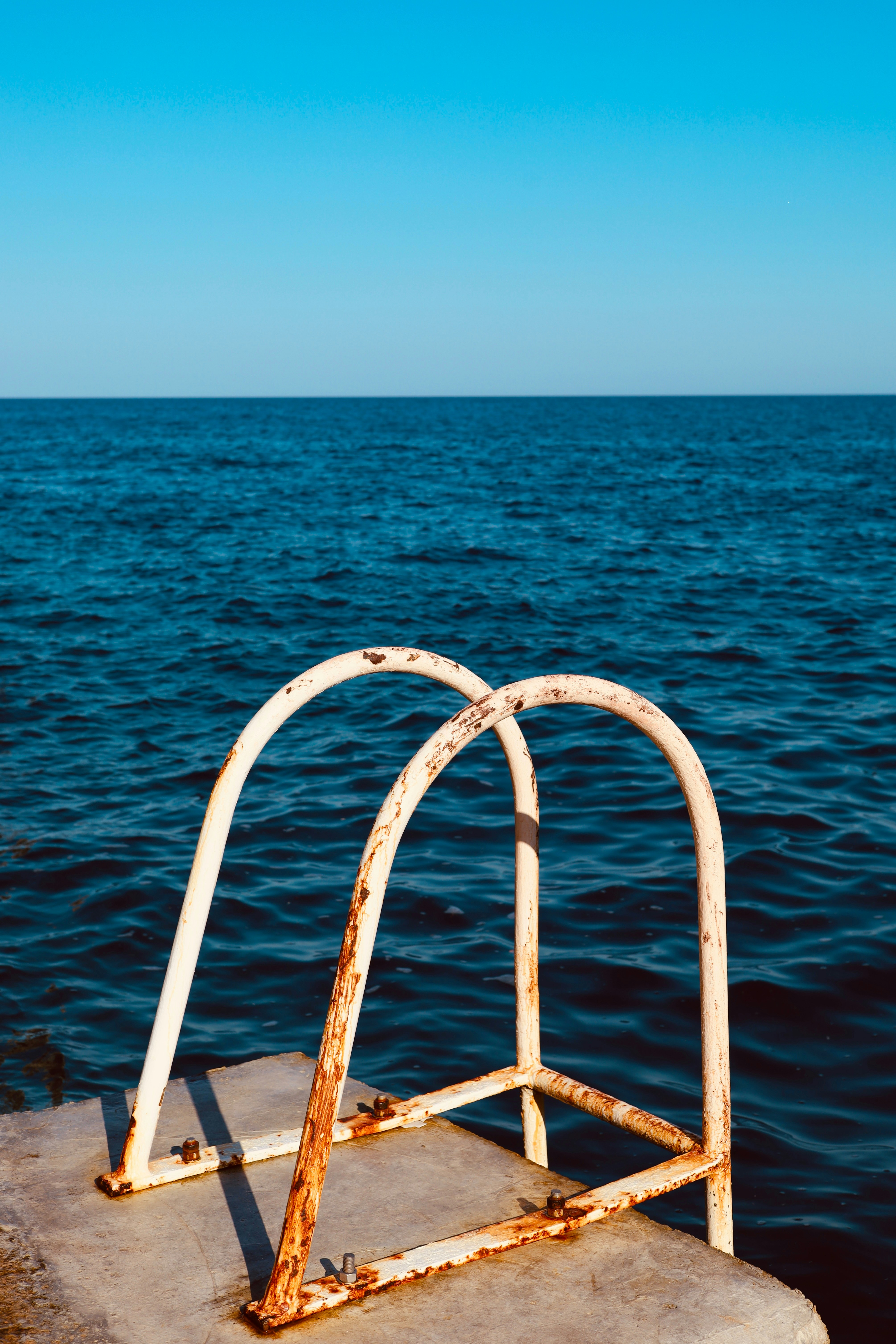 Rusty metal handrails rise from a weathered concrete platform into a calm, deep-blue sea under a clear sky.