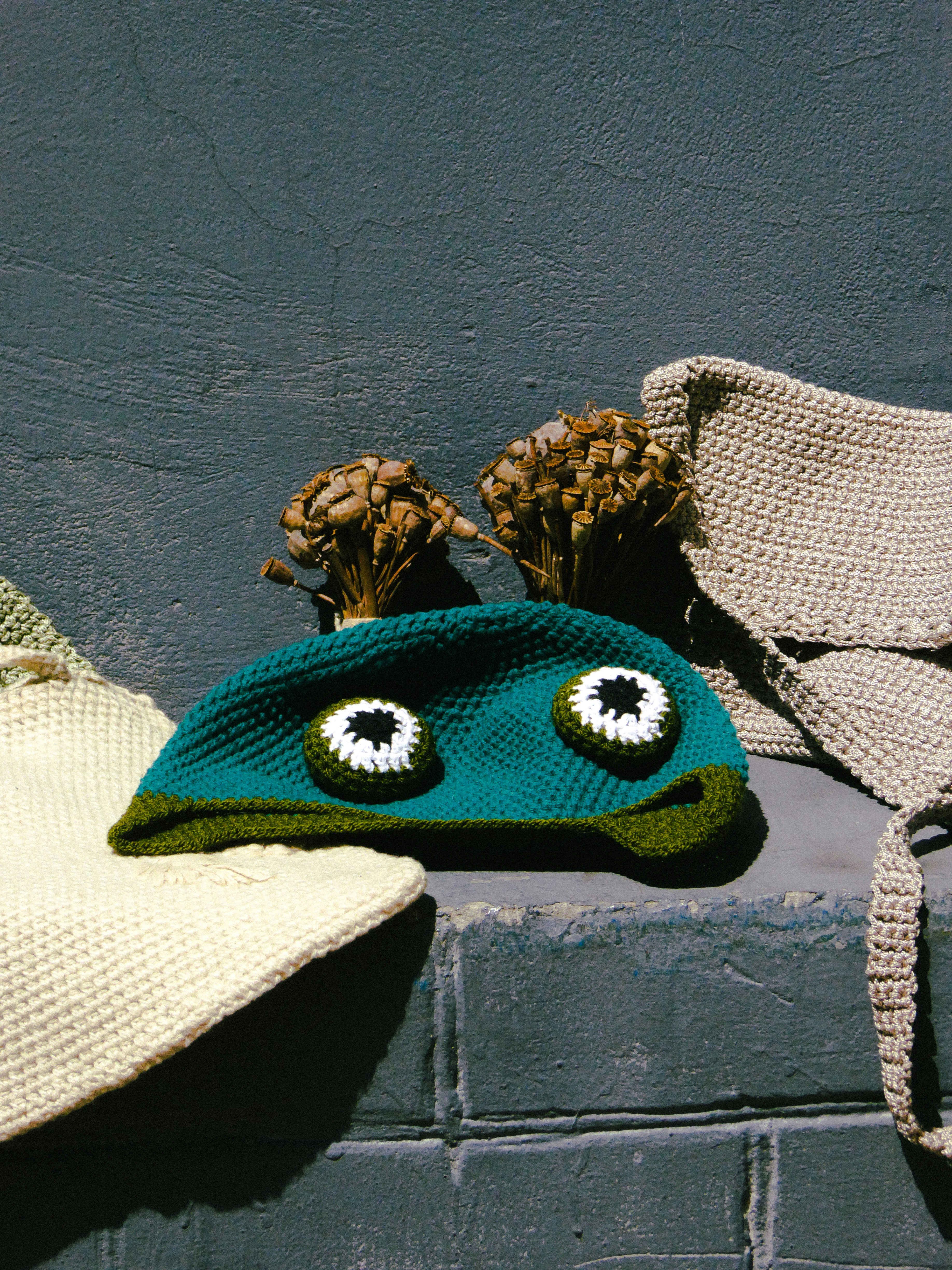 A knitted teal creature with large googly eyes rests on a concrete ledge between textured hats against a dark wall. The scene emphasizes whimsy and tactile fabric against urban backdrop.
