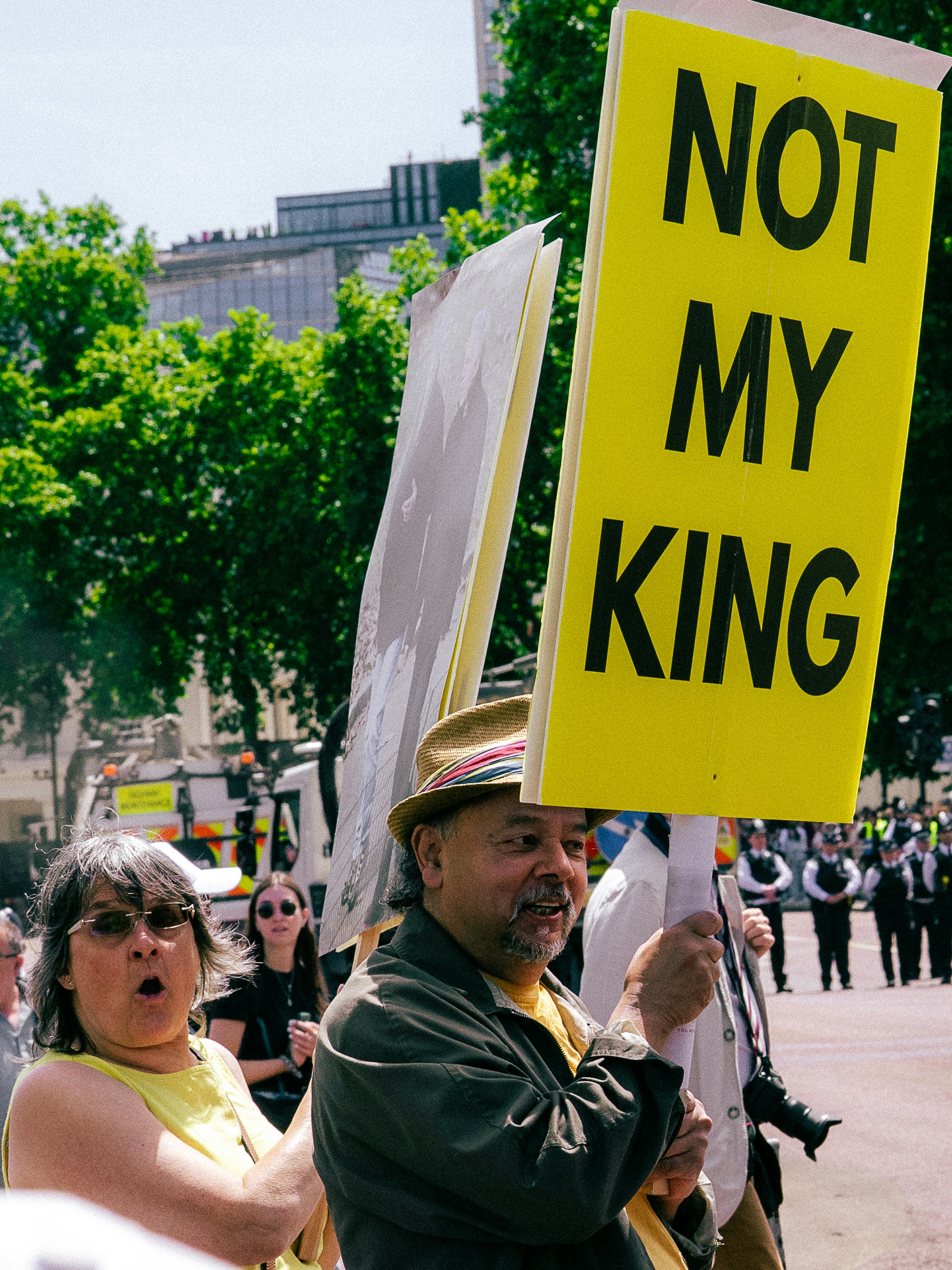Protesters in a sunlit urban square hold a bright yellow banner reading 'NOT MY KING' with onlookers and greenery in the background.