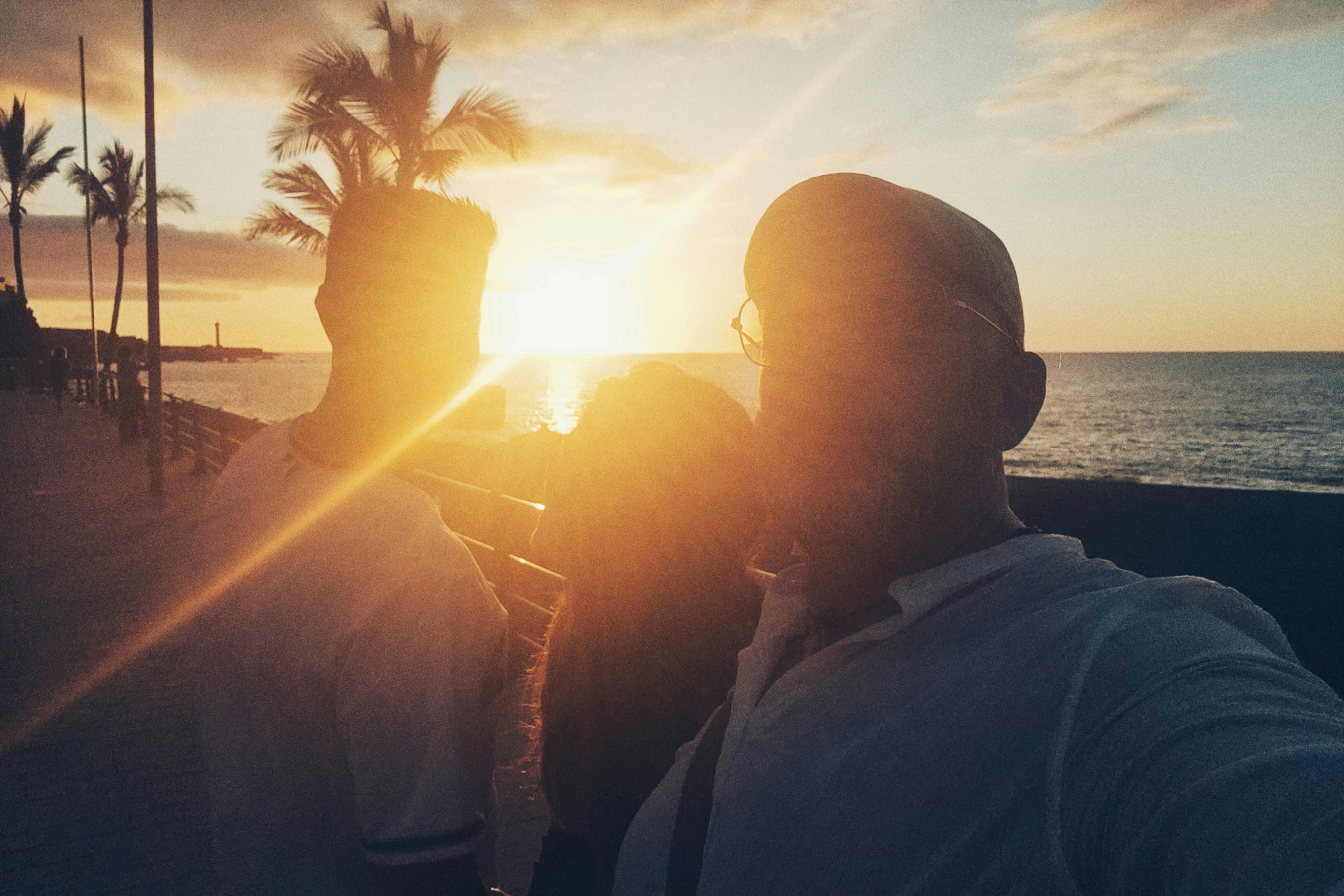 Two people silhouette against a setting sun at a tropical beach; palm trees line the left, ocean on the right, with lens flare washing over the scene.