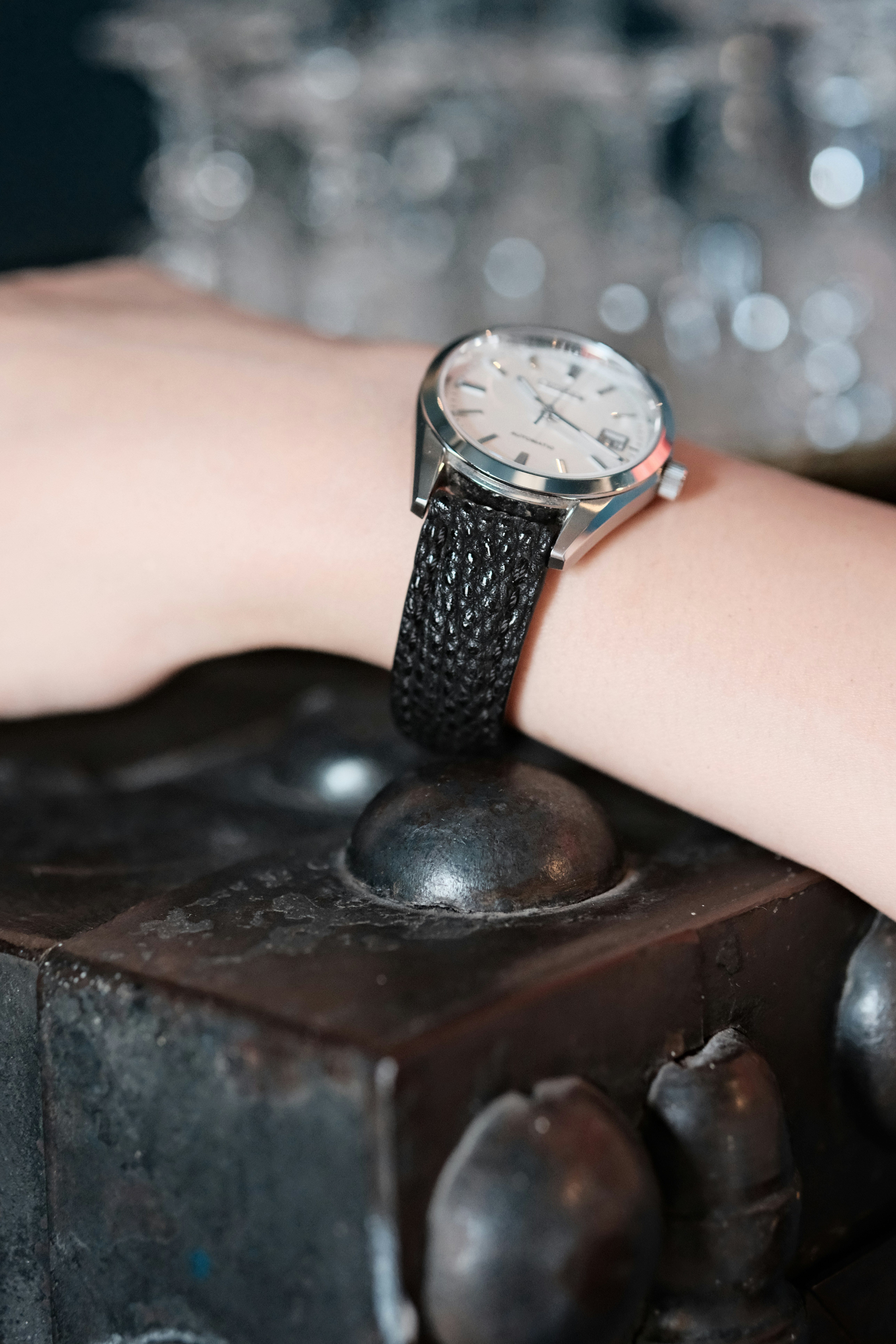 Close-up of a wristwatch with a black textured strap resting on a rounded bar knob; soft bokeh background hints at glassware. This is a lifestyle/product presentation.