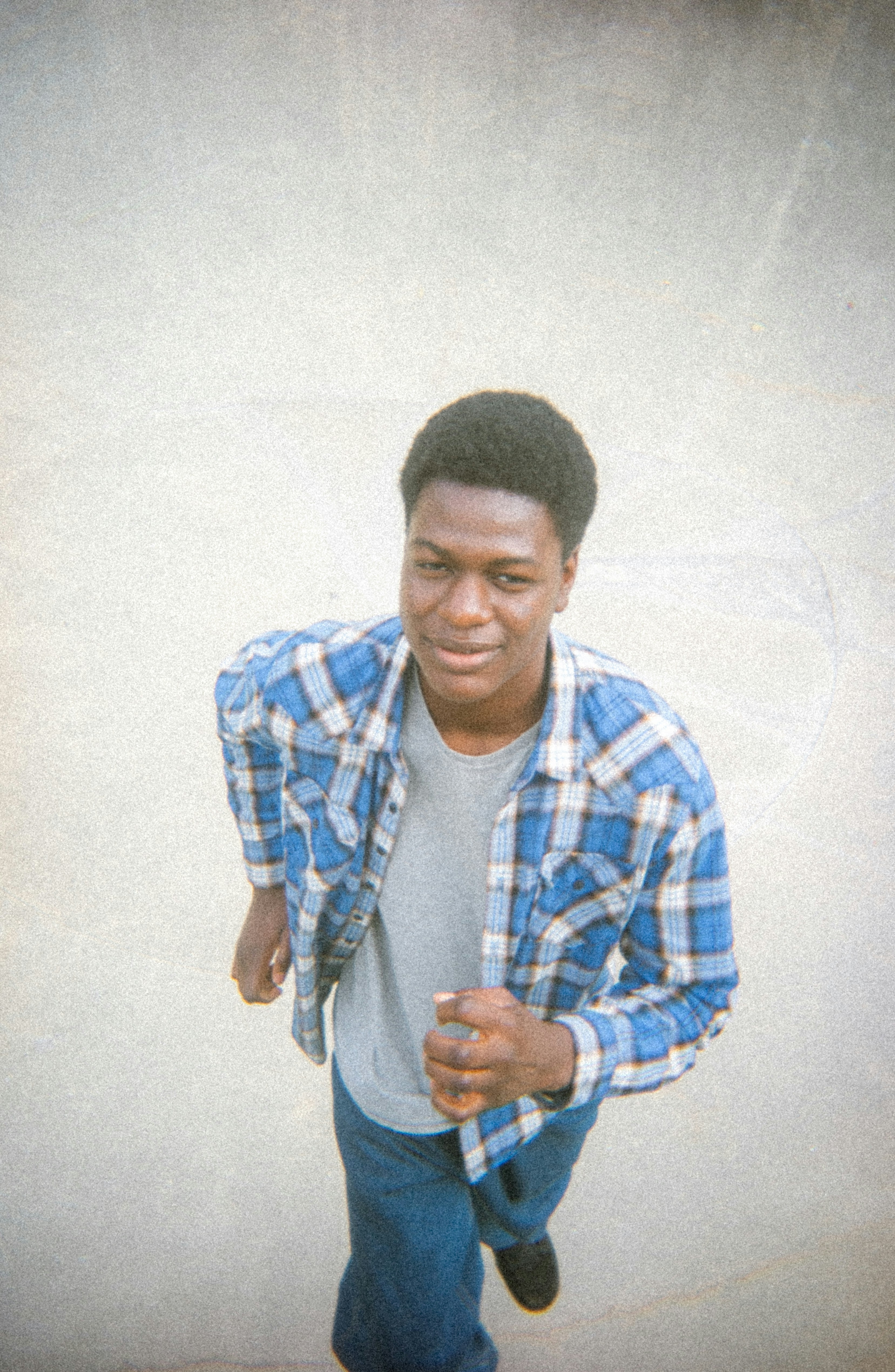 Portrait of a young man in a blue plaid shirt captured mid-step, with a soft vintage grain and muted backdrop. A casual, energetic moment emphasizing movement and expression.