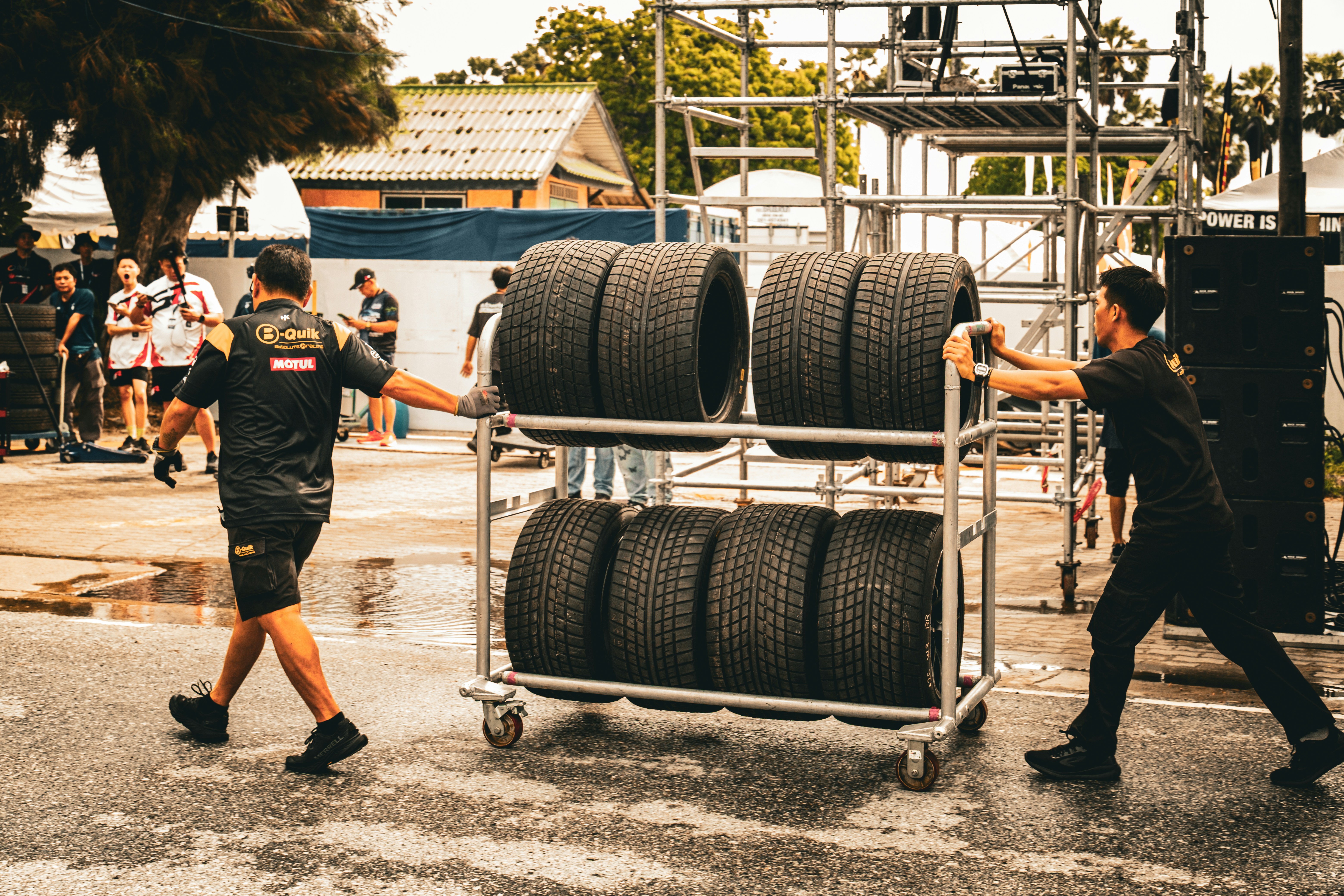 A candid behind-the-scenes documentary photograph capturing a team of motorsport paddock crew members working together to push a large heavy-duty rolling metal rack stacked with Michelin wet racing tyres. Photographed in the humid temporary pit lane environment of the Bangsaen Street Circuit during the Porsche Carrera Cup Asia and Thailand Super Series. The composition highlights the physical labor, logistics, and intense preparation of the race crew working on a wet asphalt surface, framed by temporary scaffolding structure and trackside team gear. Ideal for editorial use, sports logistics fe