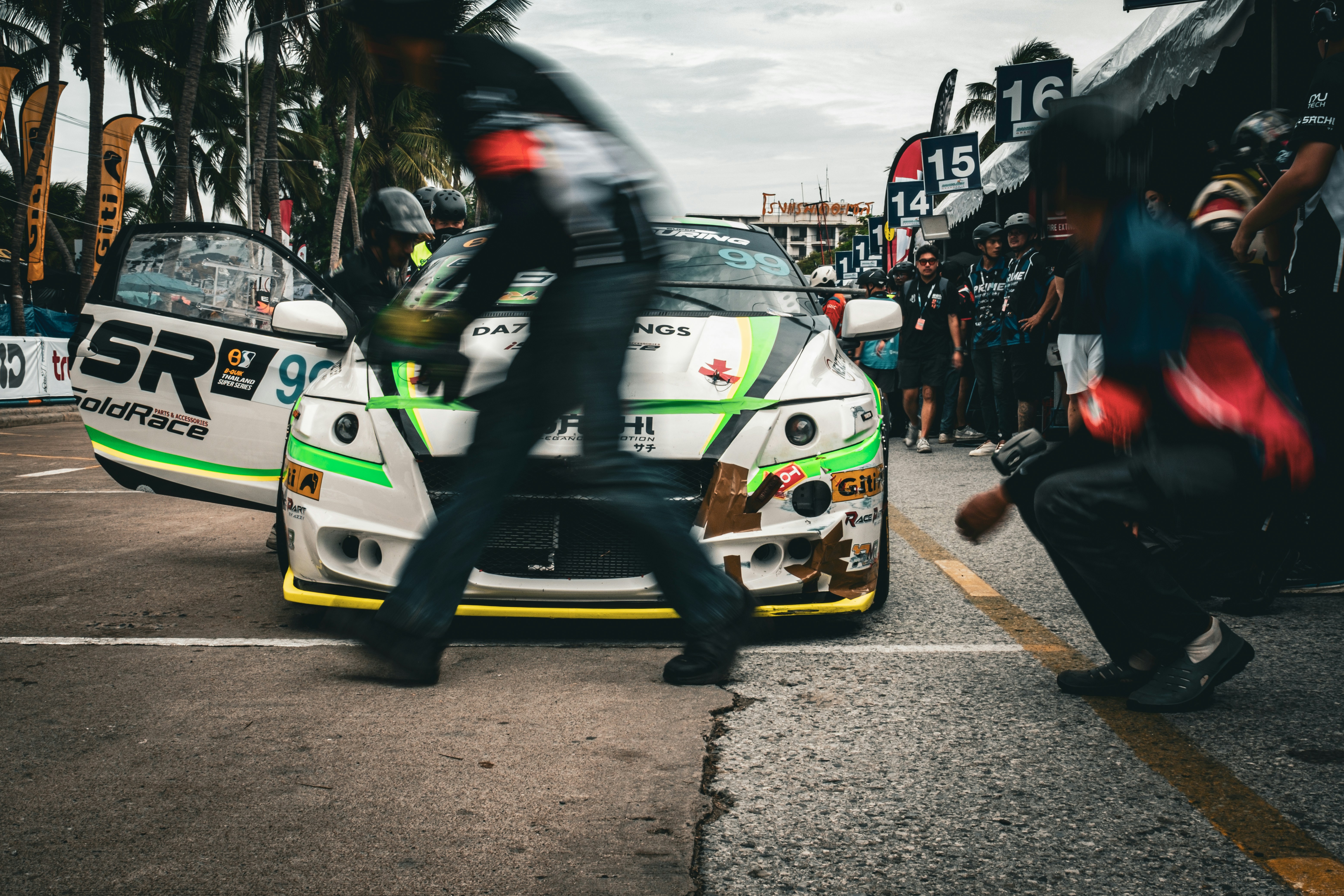 A dynamic, high-energy documentary photograph capturing the frantic atmosphere of a busy motorsport pit lane at the Bangsaen Street Circuit during the Thailand Super Series. The central focus is a white and neon-green #99 touring race car with its driver's door swung wide open, while pit crew members move in a fast motion blur across the foreground. The background showcases palm trees, team staff, and temporary pit boxes numbered 14, 15, and 16, perfectly conveying the urgency, speed, and real-time chaos of trackside team management. Ideal for editorial use, sports journalism.
