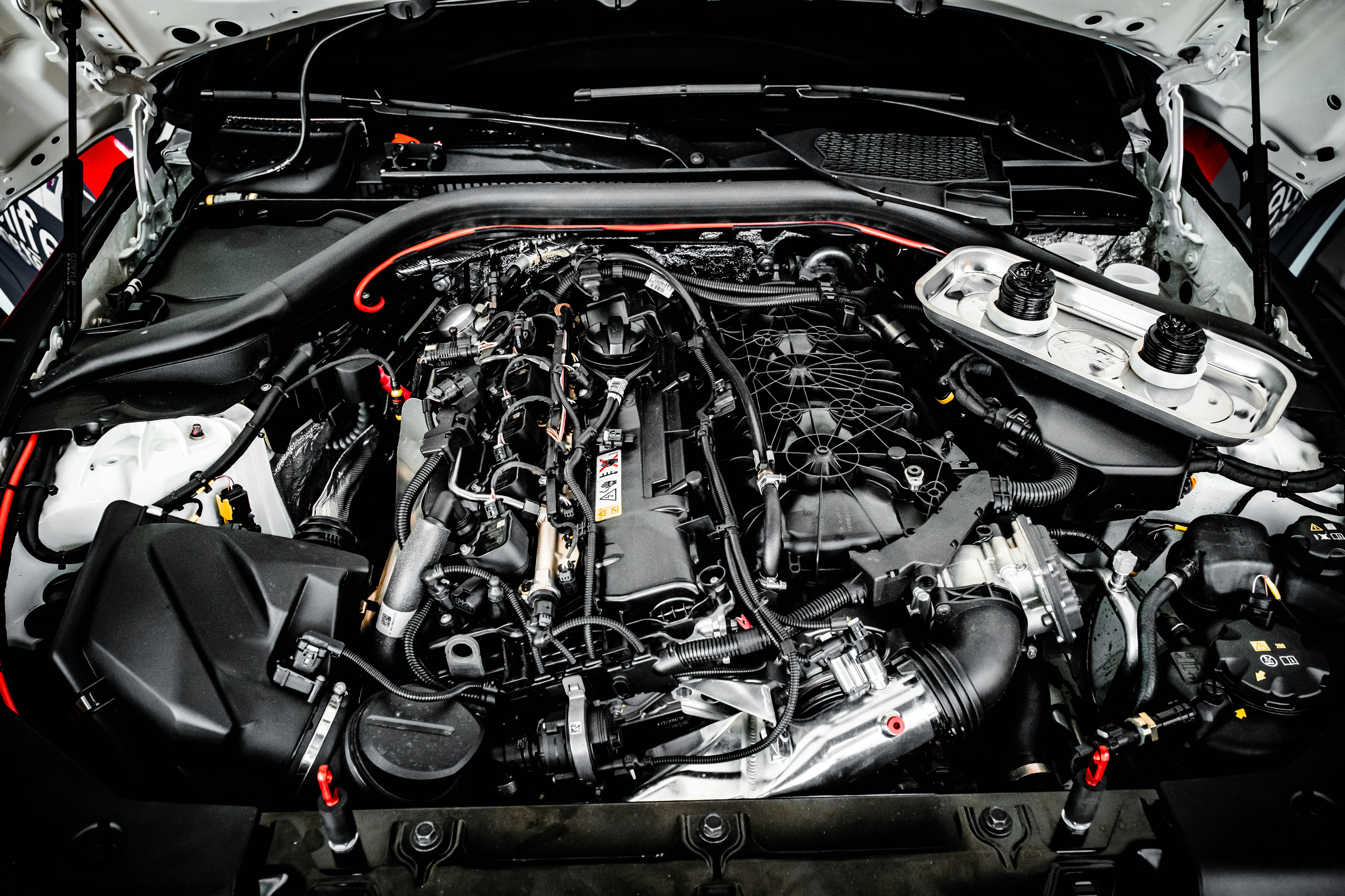 A high-angle, detailed technical photograph of an open engine bay showcasing a high-performance inline 4-cylinder turbocharged racing engine. Captured in the paddock garage during a Touring Car Asia and Thailand Super Series event weekend, the clean layout exposes a specialized composite intake manifold, intricate fuel rails, custom wiring harnesses, an aluminum turbo charge pipe, and fluid reservoirs under an open hood. This crisp, industrial automotive image is perfect for mechanical engineering articles, motorsport technical features, car maintenance guides, and high-performance aftermarket