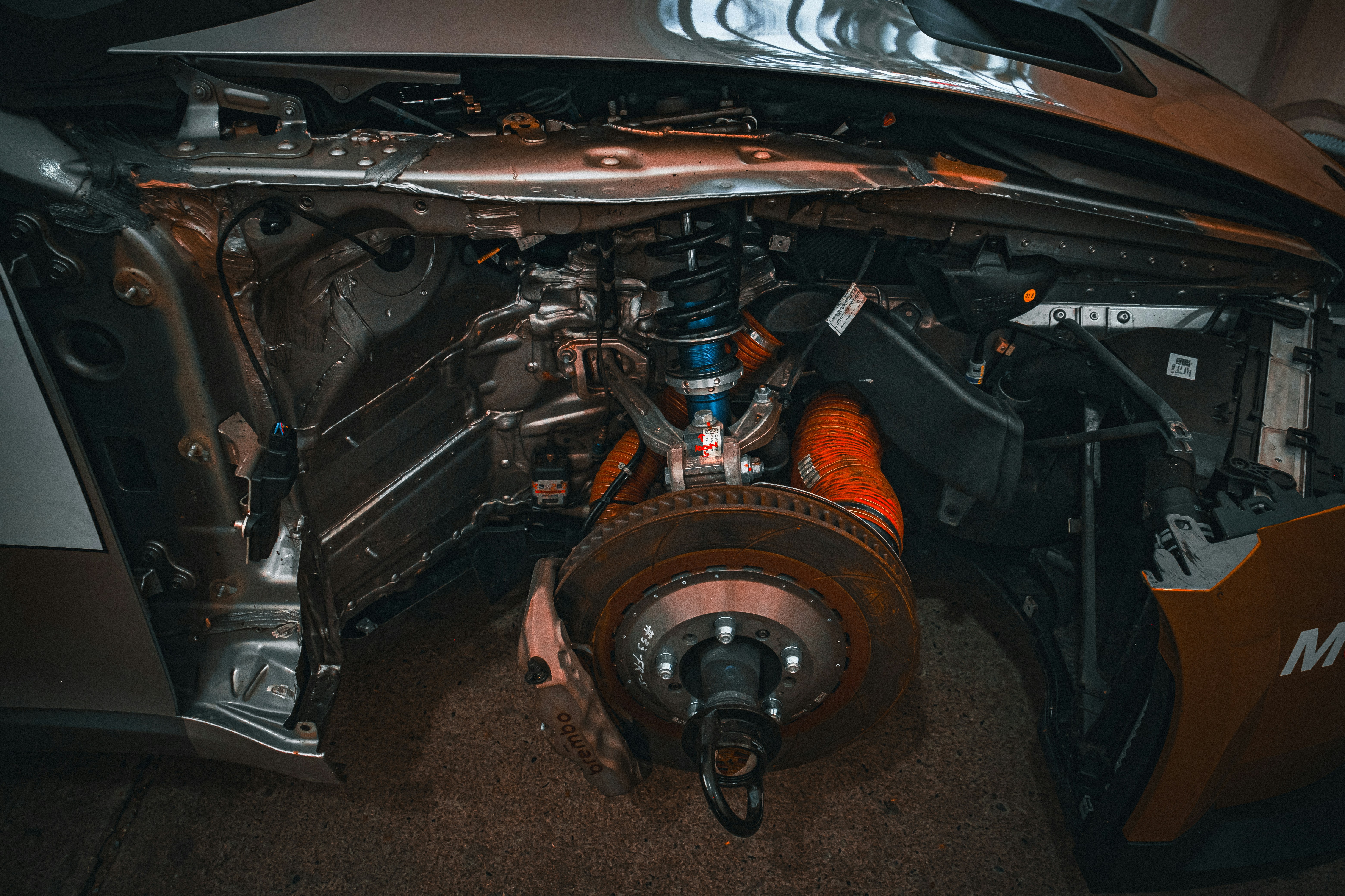 A wide, technically detailed photograph of the exposed front-wheel well assembly on a Porsche 911 GT3 Cup race car during the Porsche Carrera Cup Asia paddock preparations. With the wheel and front fender paneling removed, the image reveals a premium Brembo racing brake caliper, a slotted steel brake disc rotor, adjustable coilover suspension, and distinctive orange high-temperature brake cooling duct hoses snake-routing through the inner chassis. Ideal for editorial coverage, motorsport technical analysis, high-performance automotive blogs, and industrial engineering stock photography.