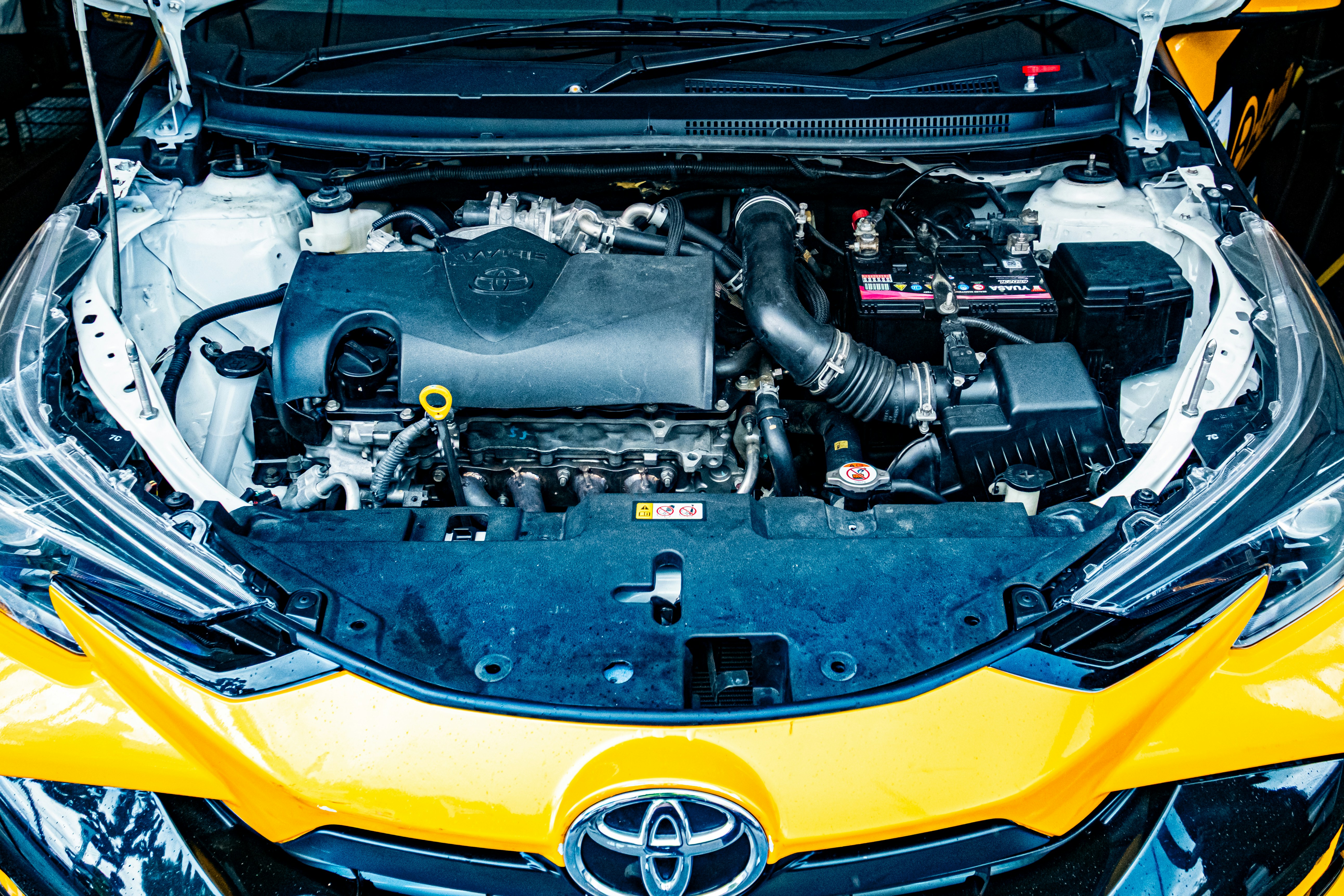 Top-down wide photograph of an open engine bay showcasing a modern Toyota Dual VVT-i 4-cylinder engine. Documented in the paddock during the Touring Car Asia event weekend, the clear view reveals the engine cover, air intake duct, Yuasa car battery, and fluid reservoirs framed by the bright yellow front bumper. Excellent for automotive maintenance blogs, car repair guides, and technical stock imagery.