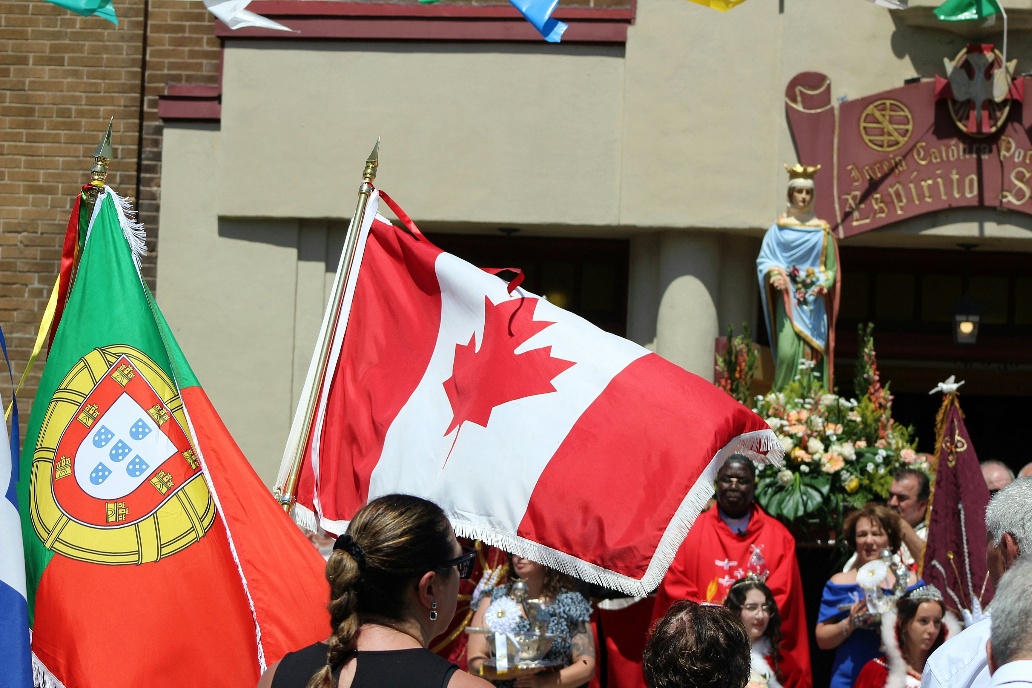 A crowd gathers with multiple flags, including a prominent Canadian flag, during a festive outdoor event; people and flags fill the frame with colors and celebratory banners visible in the background.