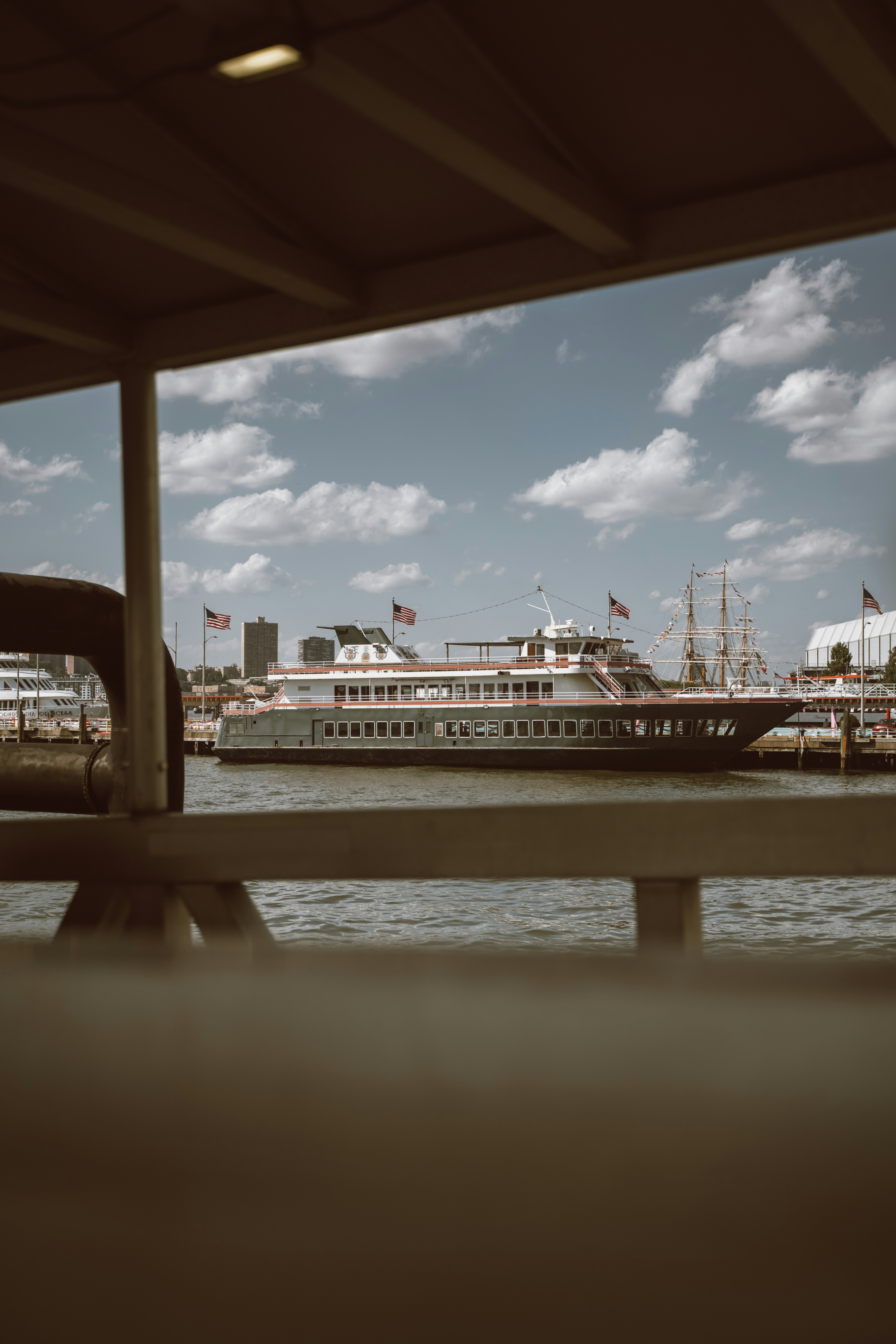 A passenger boat rests at a harbor, viewed through a shaded foreground railing and beams with a clear sky and distant masts in the background.