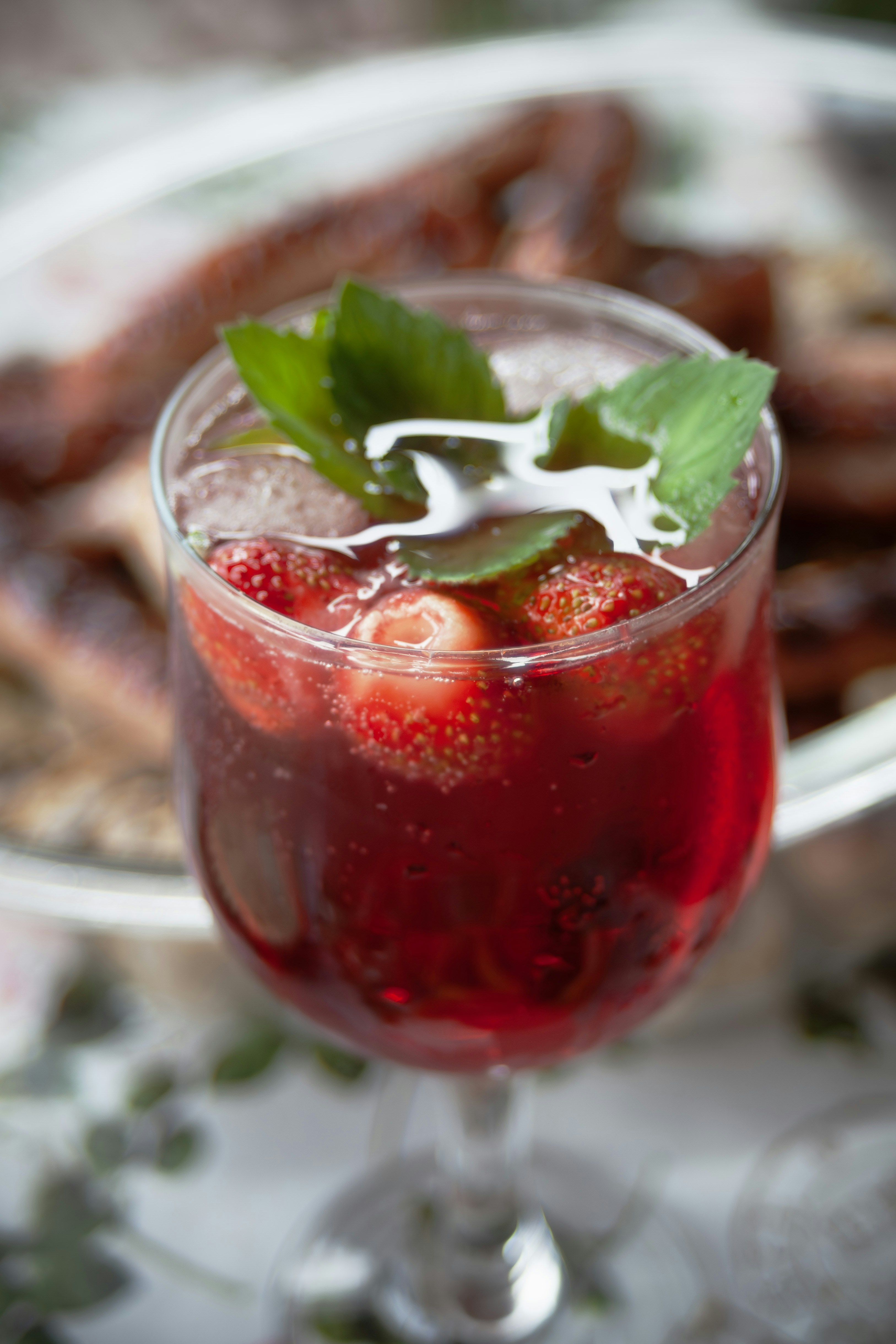 A glass of strawberry spritzer with mint leaves sits in foreground, blurred dish in background; a lifestyle close-up emphasizing the fresh fruit and beverage presentation.