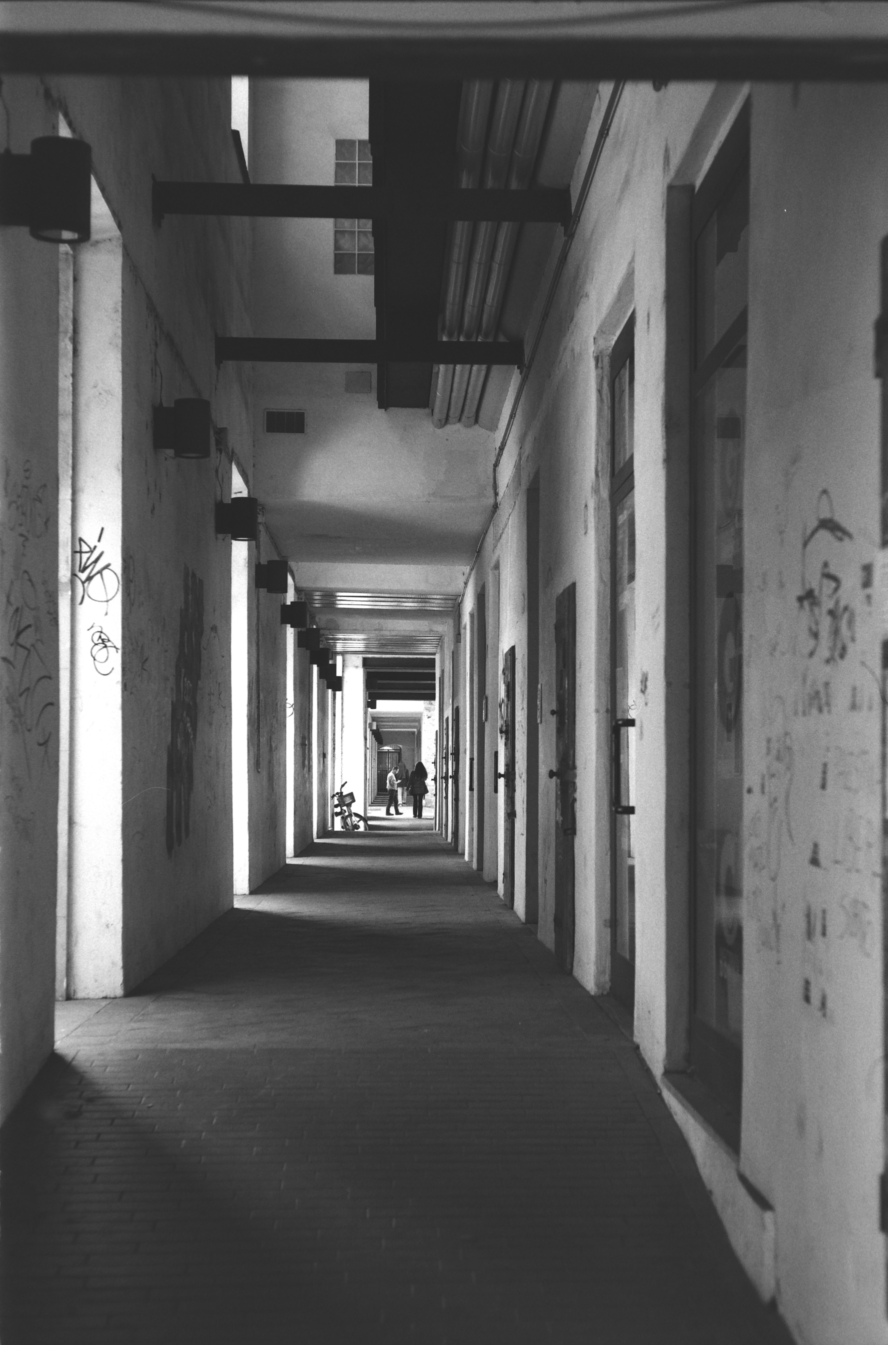Monochrome corridor with repetitive arches and distant figures, hinting at a quiet moment of passage and anonymity within an urban space.