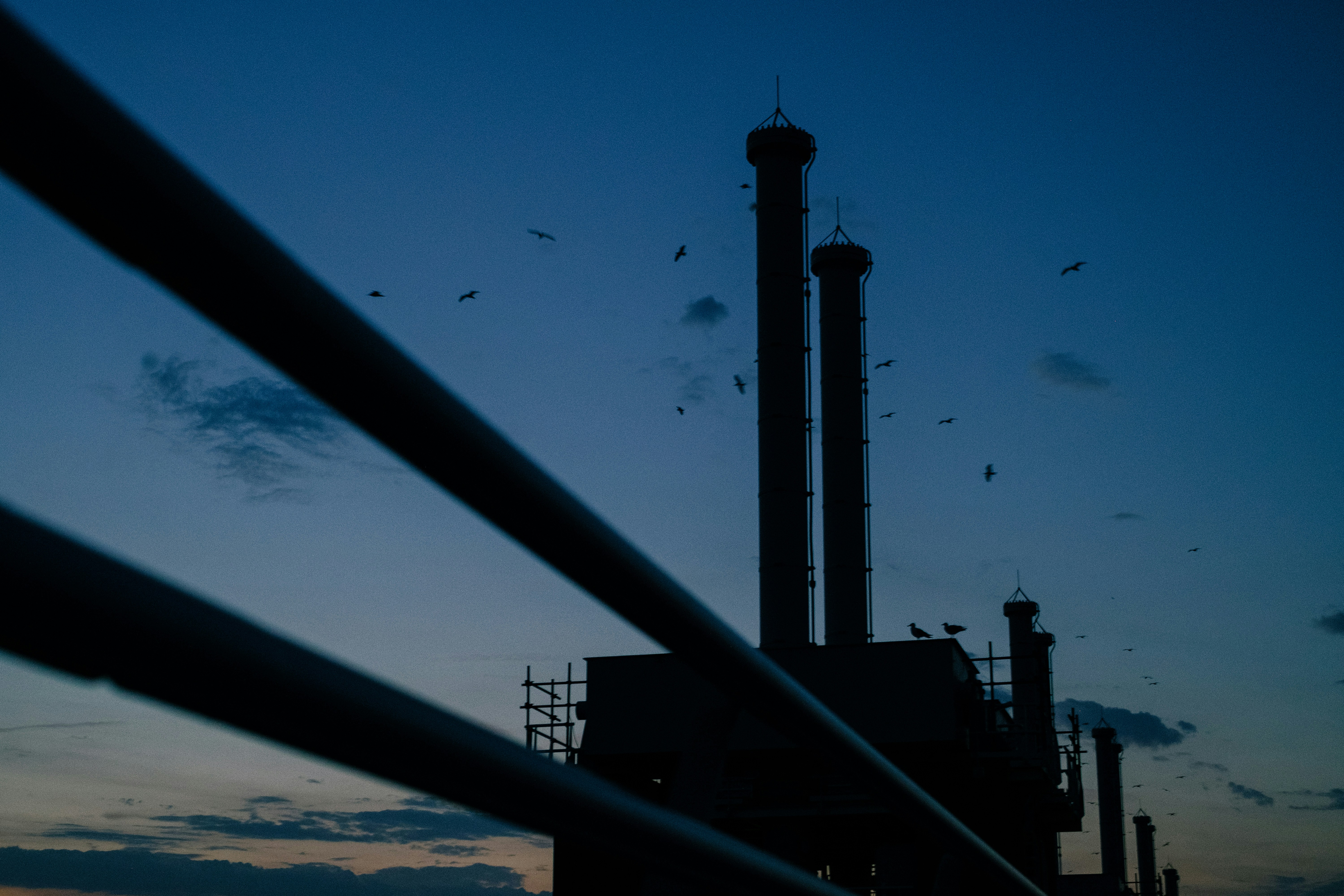A cinematic, silhouetted view of the towering structures of the Oosterscheldekering storm surge barrier at dusk. A flock of birds circles the industrial pillars against a deep blue, grainy twilight sky, while blurred diagonal railings in the foreground draw the eye and add depth to the moody scene.