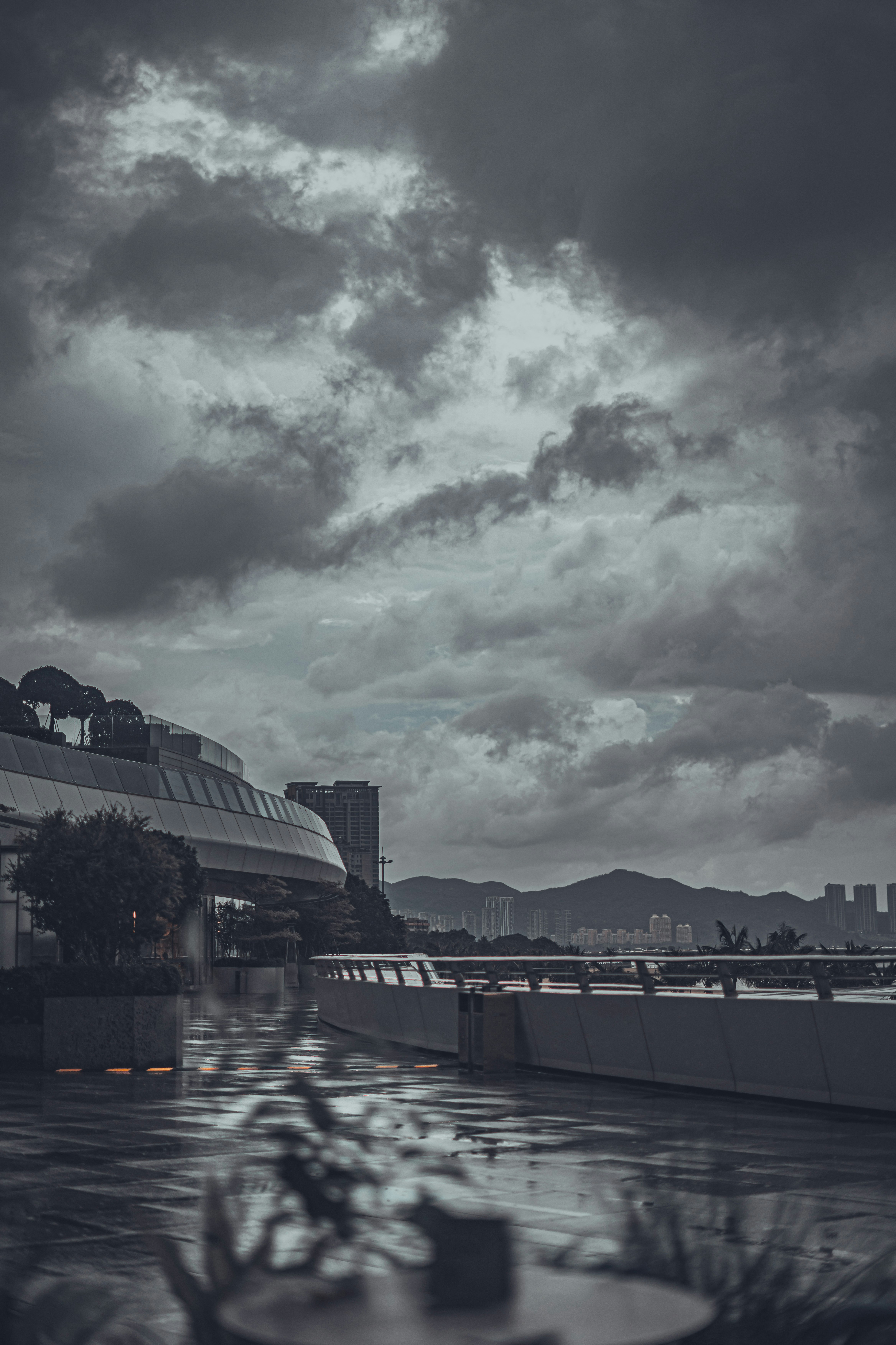 A moody cityscape along a reflective waterfront boardwalk with modern architecture and distant skyline under heavy clouds. The scene emphasizes atmosphere and architecture rather than people.