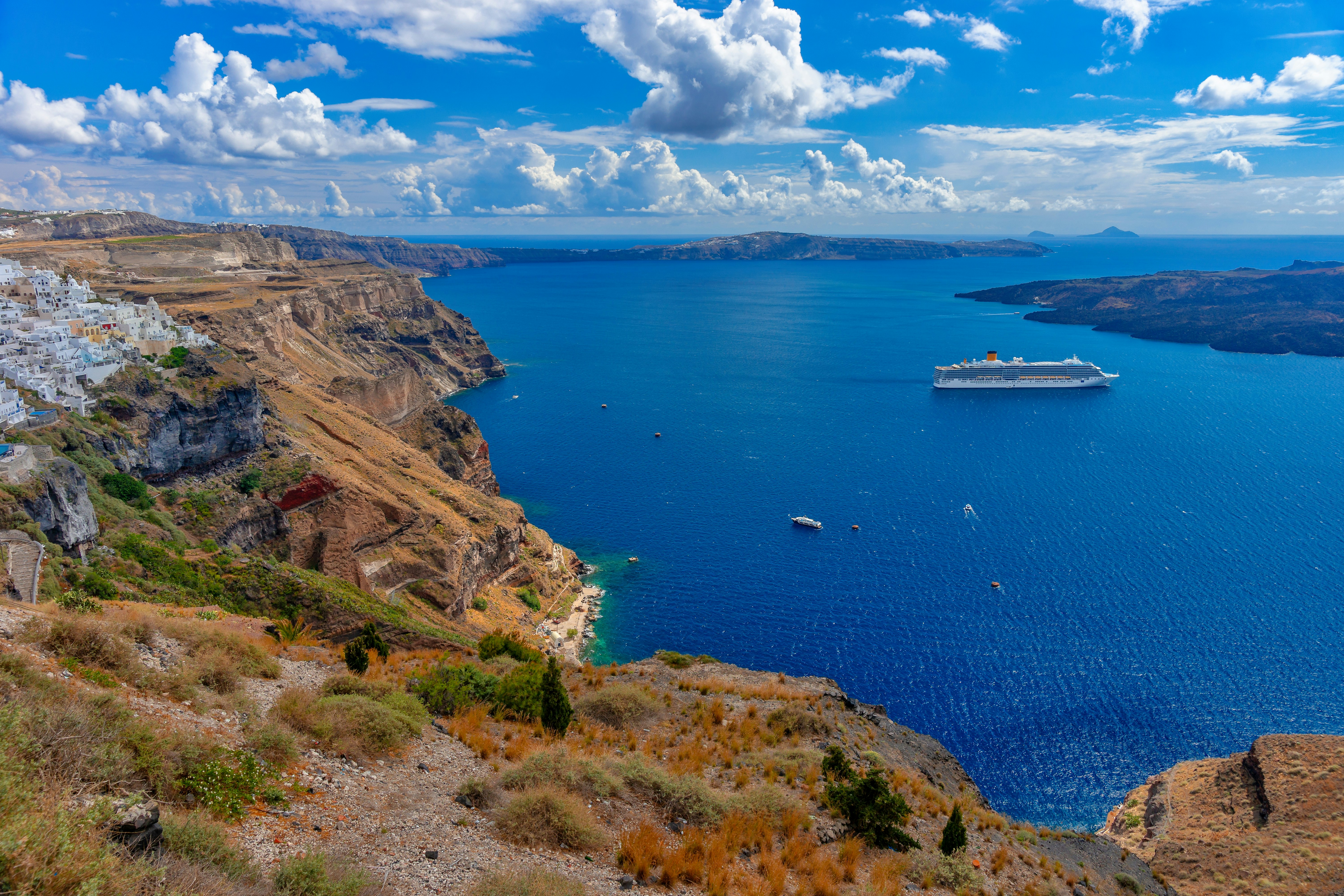 A vibrant coastal scene with rugged cliffs and a calm blue sea, featuring a large cruise ship anchored near the shore. The image emphasizes expansive seascape and distant horizons with clear skies.