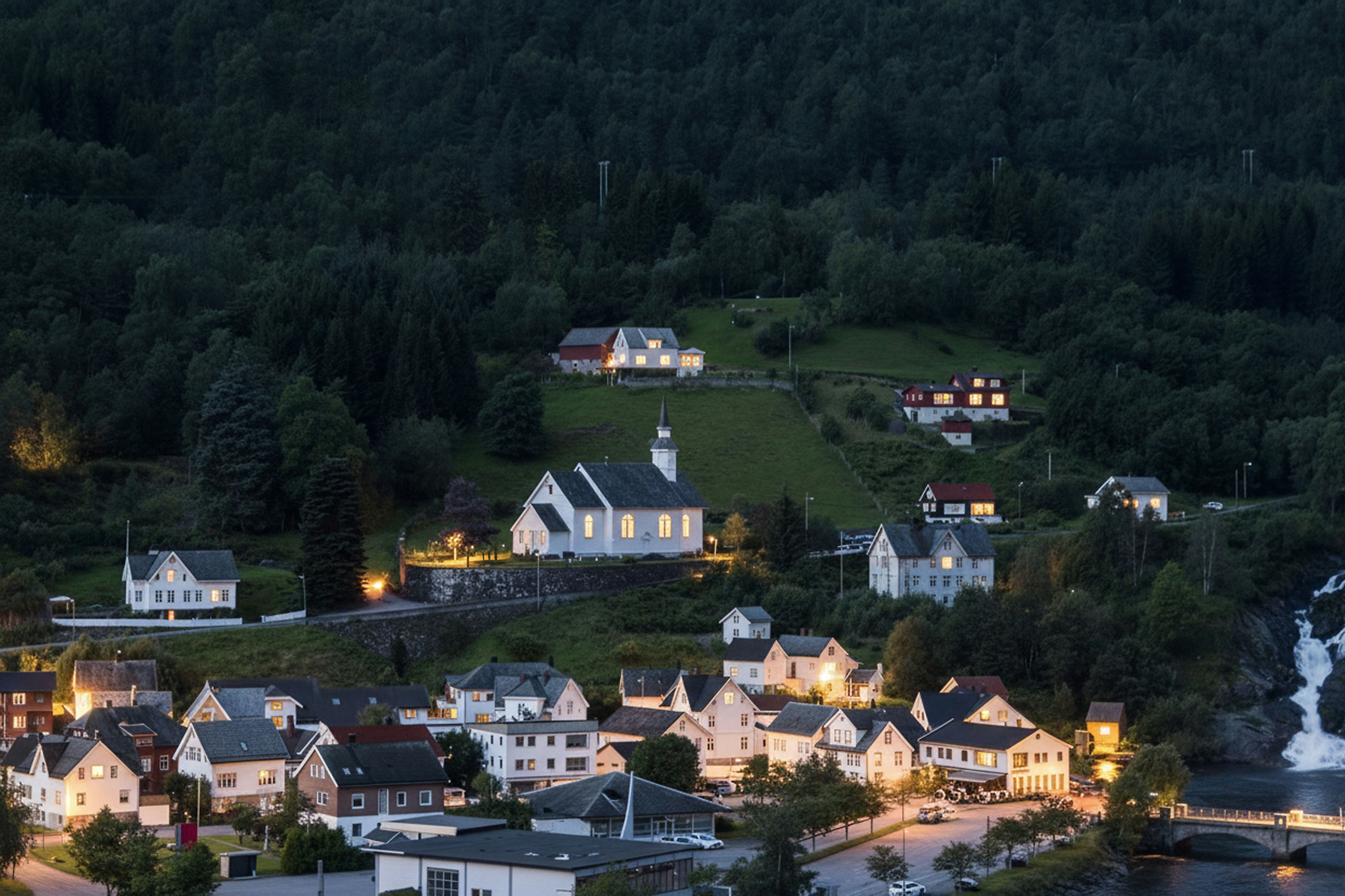 A peaceful evening view of Hellesylt, a charming village in western Norway, featuring its iconic white church illuminated at twilight, traditional Scandinavian homes, a cascading waterfall, and lush forested mountains. This picturesque fjord village offers an authentic Nordic atmosphere and serves as a gateway to the spectacular Geirangerfjord.