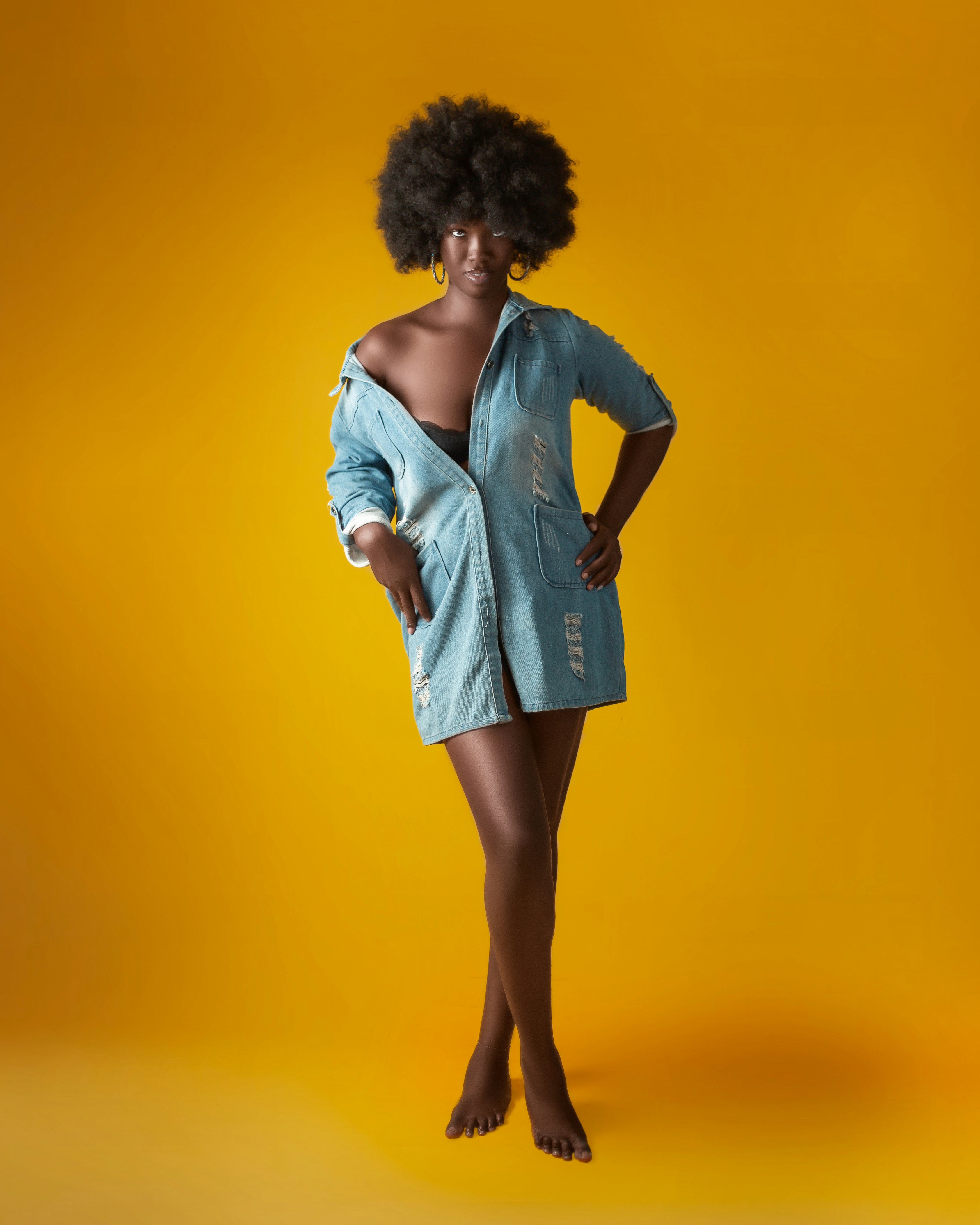 Tall model in a distressed denim shirt-dress stands barefoot against a warm yellow backdrop, posing with one hand on hip and the other gesturing forward. The scene emphasizes vibrant color and confident stance.