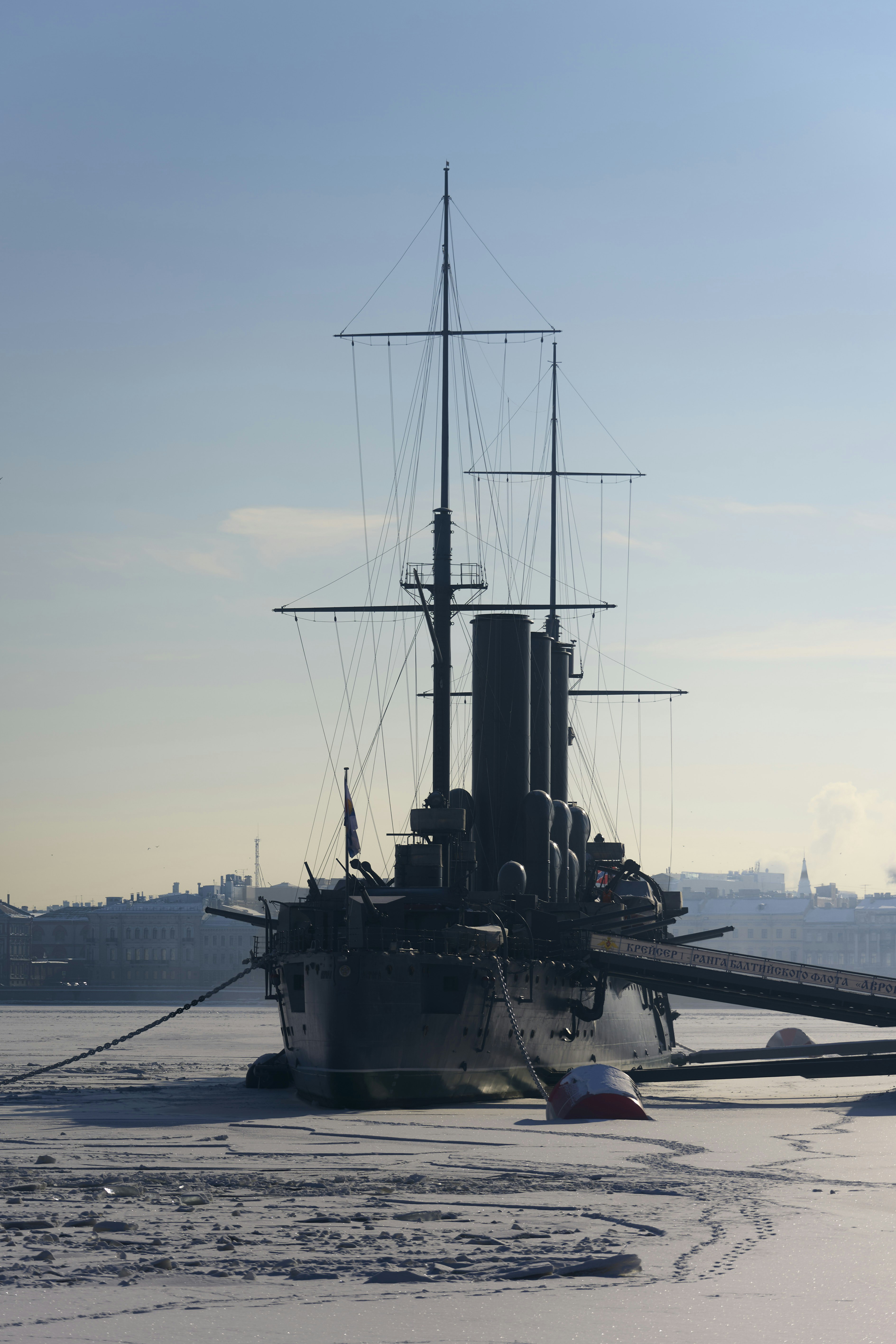 Dark battleship rests on a flat, icy shoreline with masts and rigging under a clear sky. A distant city skyline anchors the horizon.