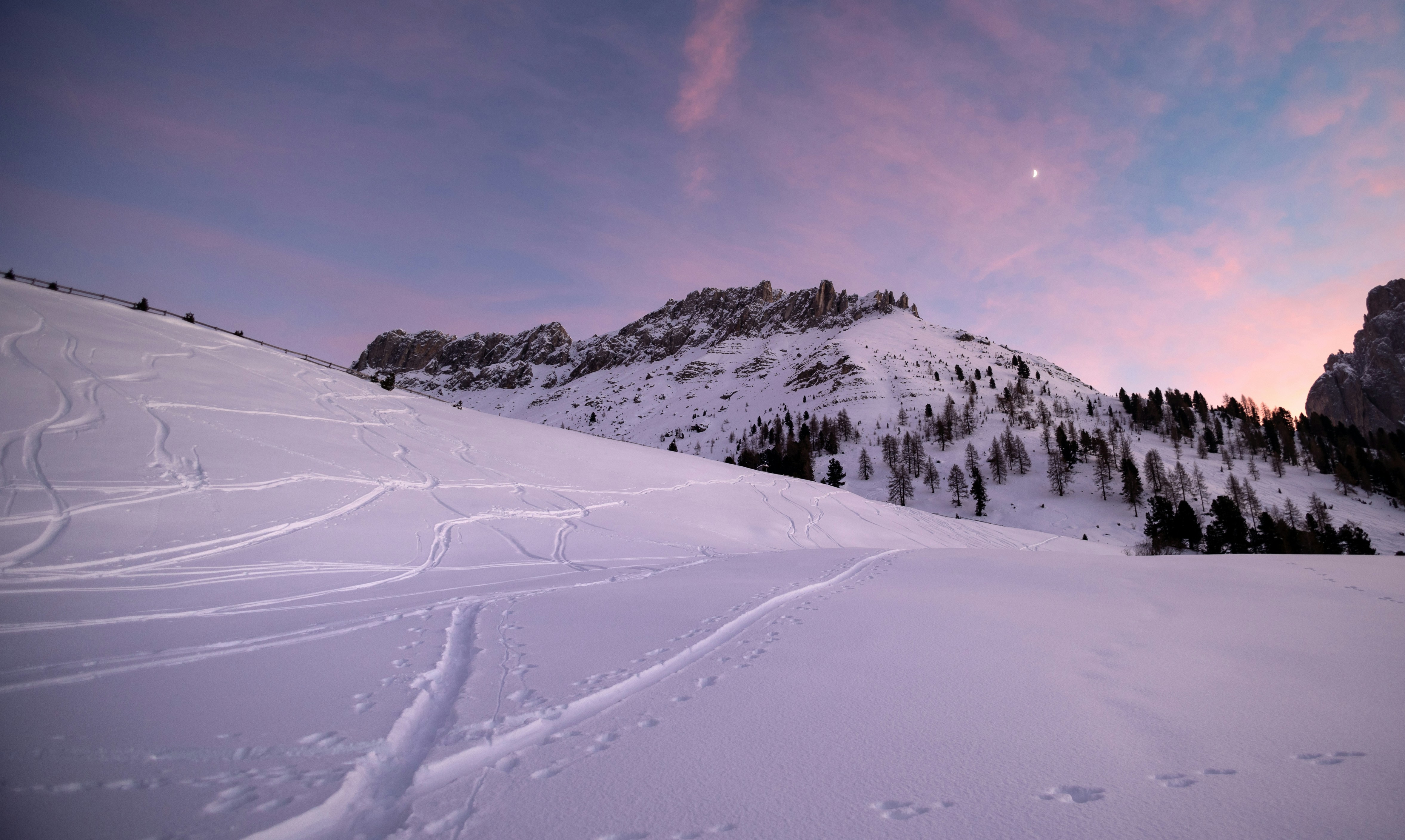 Snow-covered slope with skier tracks leading toward a jagged ridge under a pink-tinted sky. A distant hillside and trees anchor the landscape; the scene emphasizes quiet elevation and winter silence.