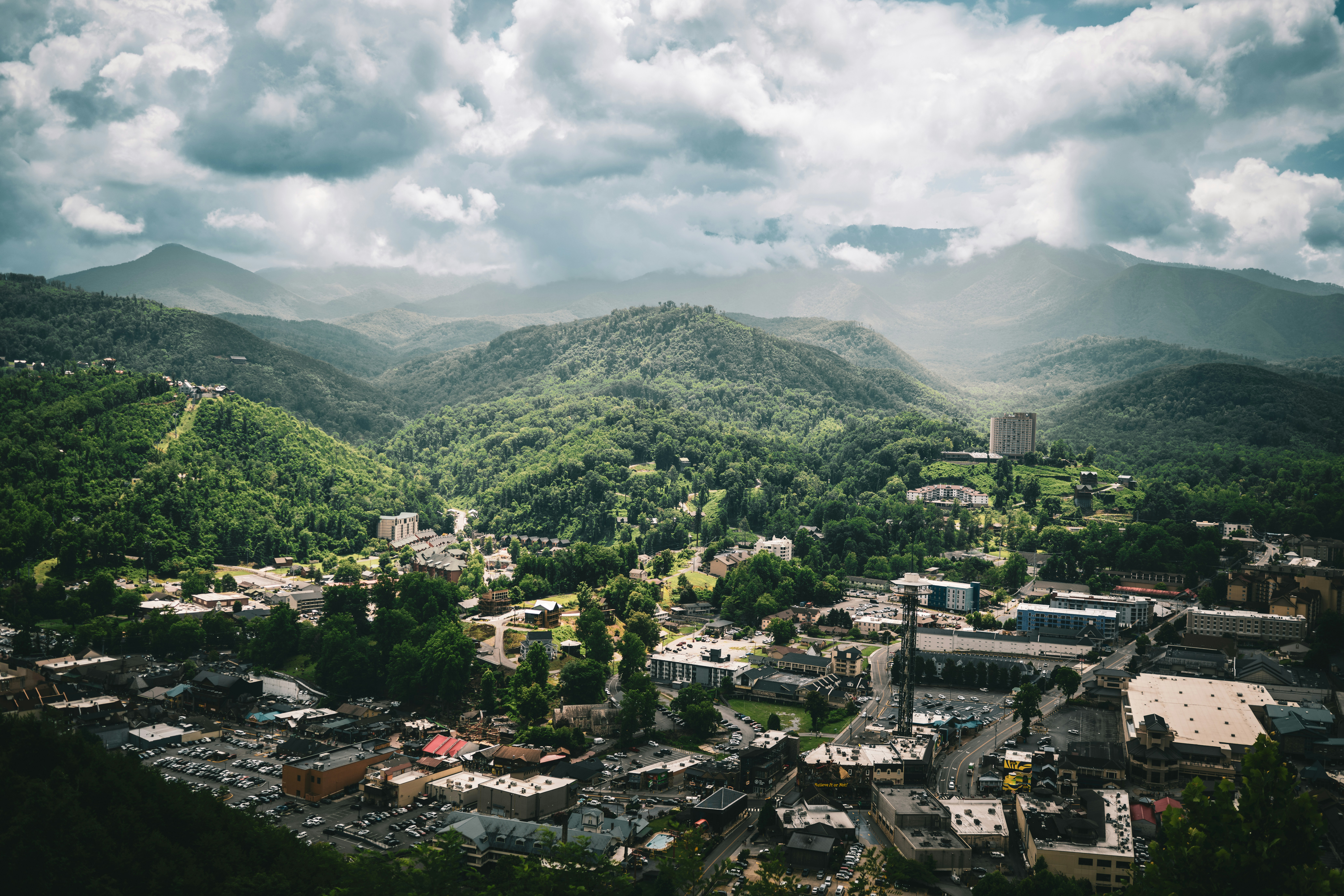 A valley town sprawls along a river with green hills and distant mountains under dramatic clouds. The scene emphasizes urban textures contrasting with natural slopes; landscape shot capturing scale and depth.