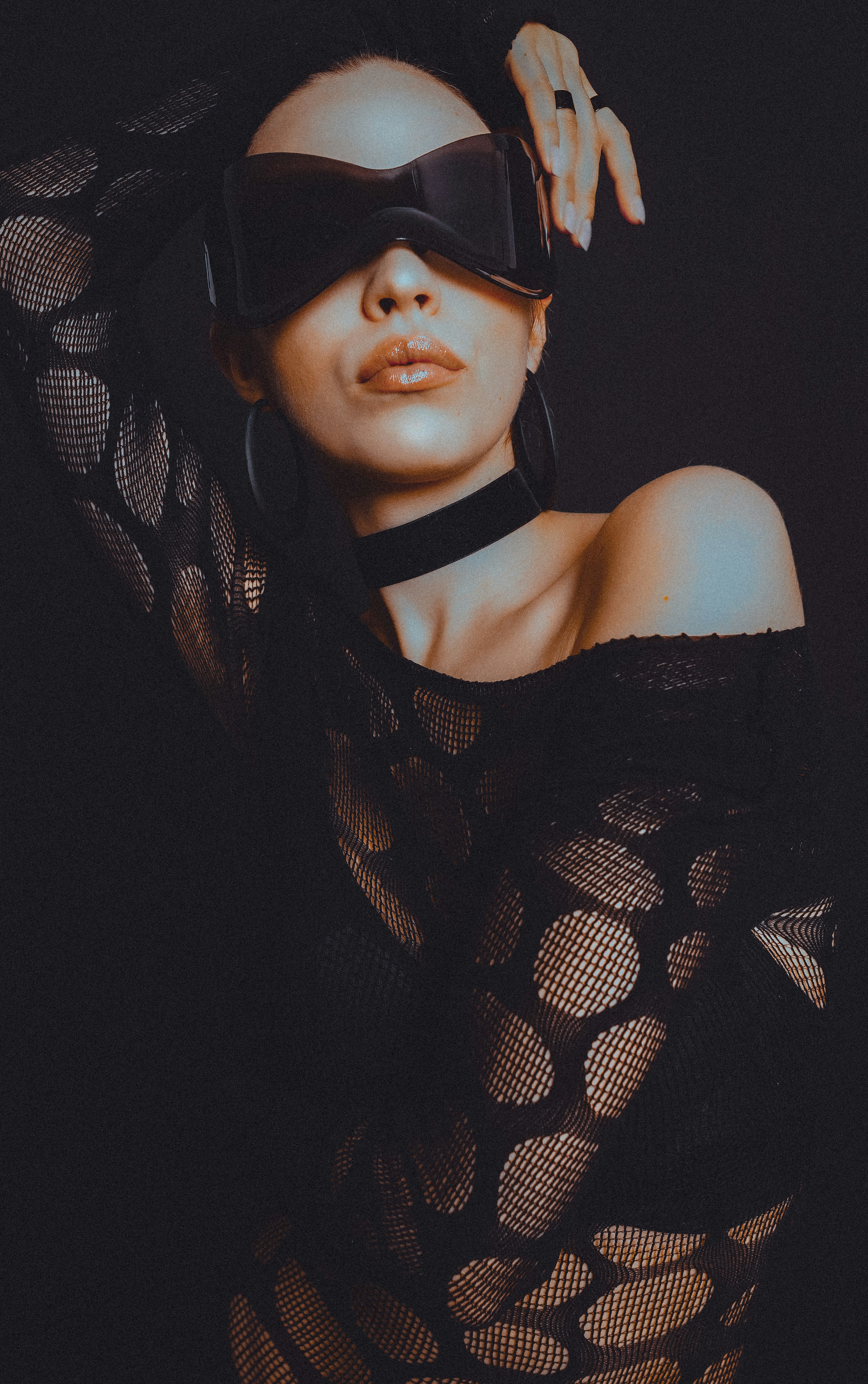 Close-up portrait of a woman wearing avant-garde black mask and netted garment, posing with a sensuous vibe in a dark studio setting.