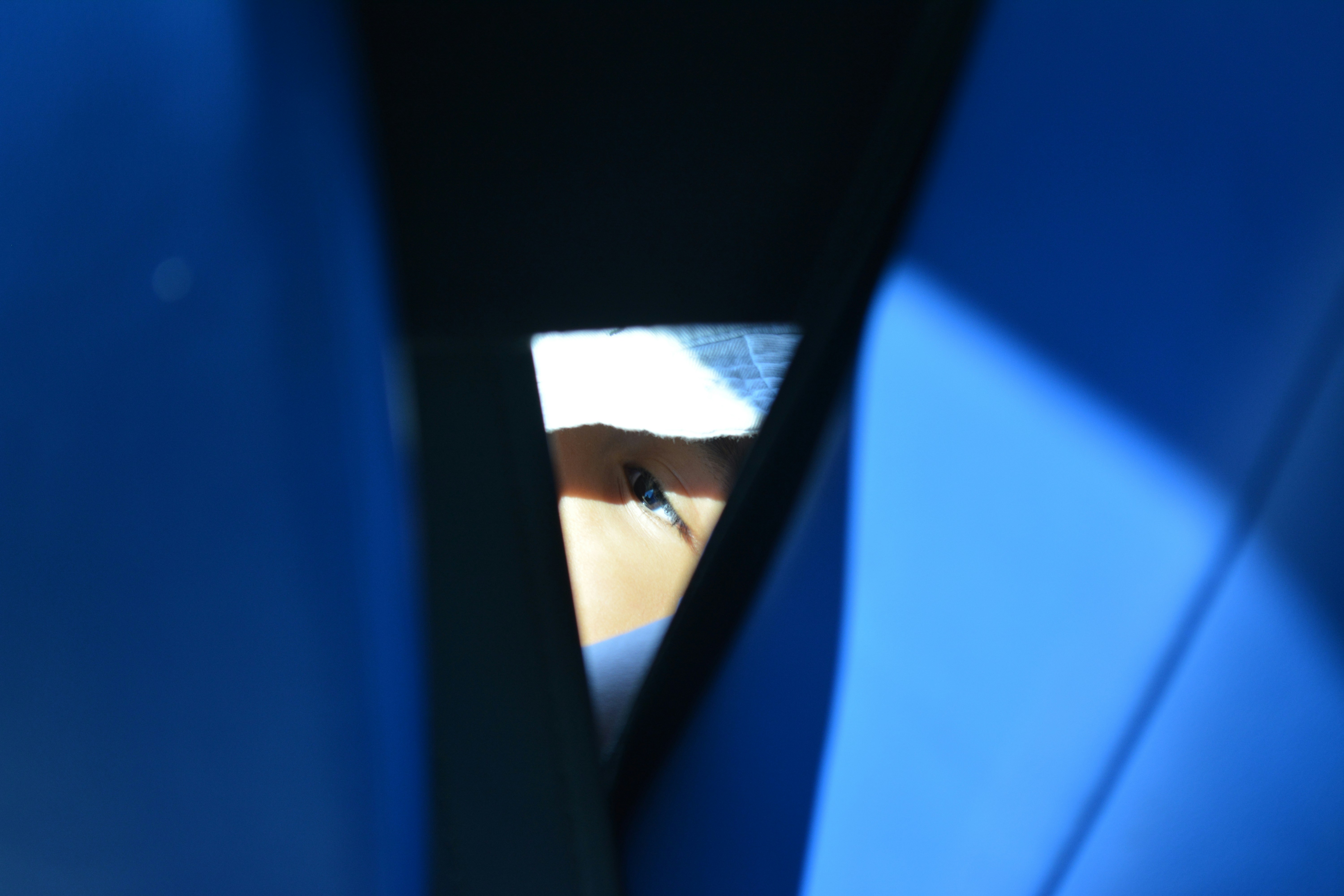 An abstract, high-contrast composition framing a child's eye looking through a narrow, triangular opening. The intense blue geometric shapes in the foreground and a stark black shadow form a tight vignette, drawing complete focus to the subject's soft gaze. Catching a sliver of bright daylight, the eye reflects a subtle glint of curiosity. This artistic, minimalist photograph masterfully uses negative space, geometric angles, and dramatic lighting to create a powerful sense of mystery, peeking, and innocent childhood observation.