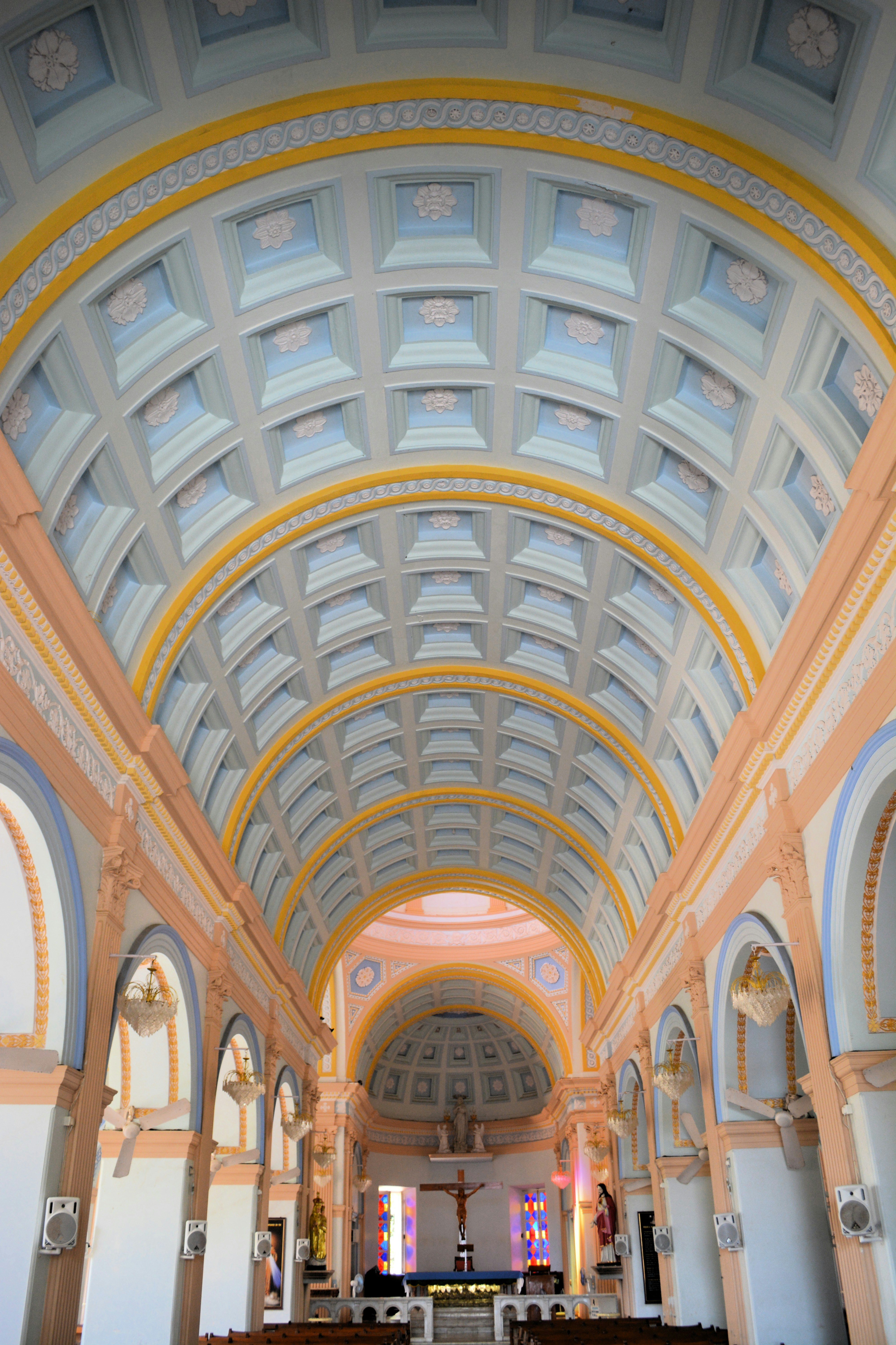 A spectacular low-angle view looking down the nave toward the altar of Our Lady of Angels Church (Église de Notre-Dame des Anges) in Puducherry, India. The architectural highlight is the magnificent, barrel-vaulted ceiling painted in soft blue, yellow, and pastel peach. Deeply moulded, recessed coffers decorated with stylised rosettes line the vault arches. Crystal chandeliers hang elegantly from the columns on either side, framing the distant altar and stained-glass windows under a beautifully detailed interior dome, capturing the unique French colonial Greco-Roman architecture of White Town.