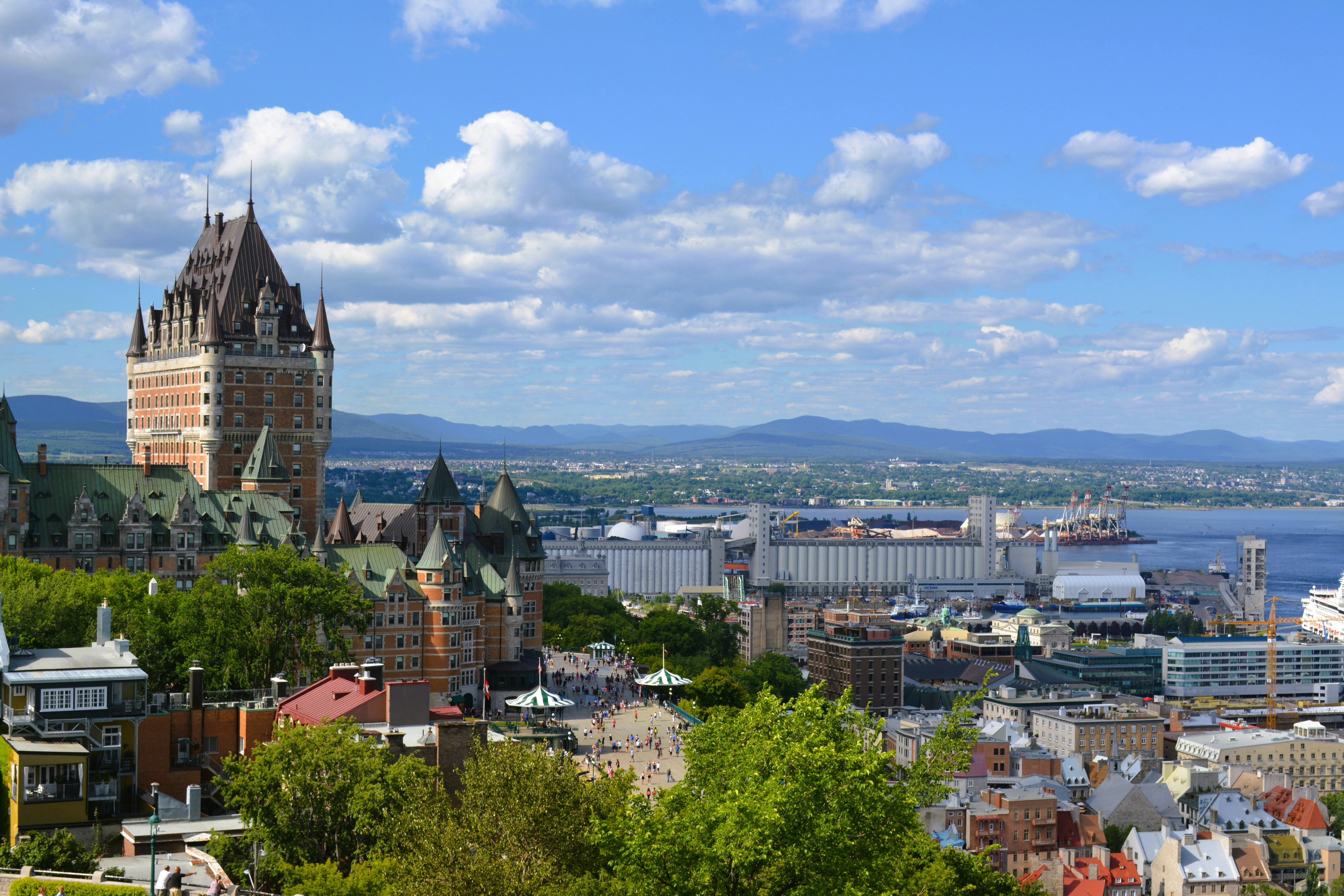 Sunlit harbor cityscape with a prominent castle-like building on the left and a mix of historic and modern rooftops under a blue, cloud-filled sky.