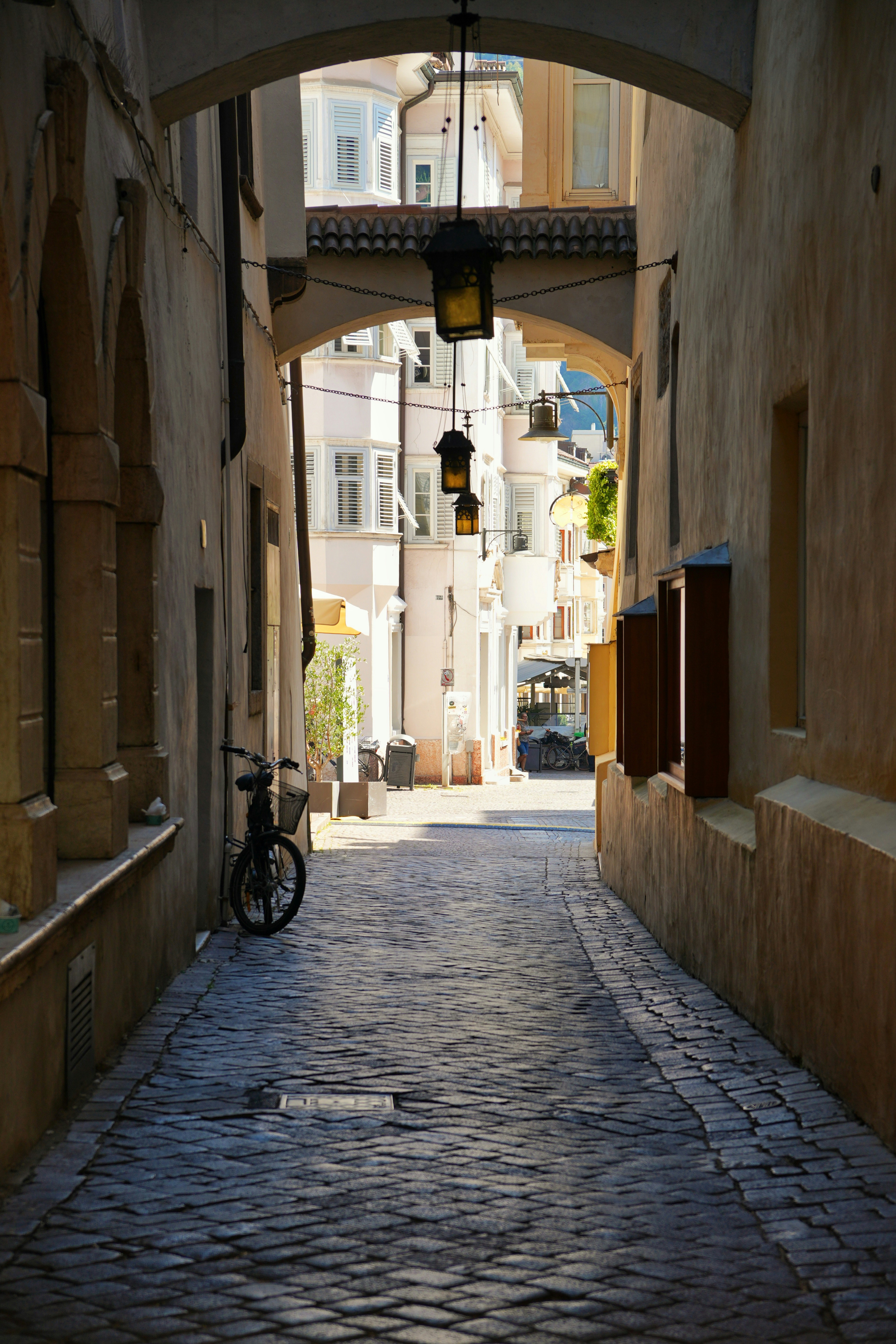 Narrow cobblestone alley framed by warm-washed walls leads to a sunlit courtyard, lanterns suspended above and a bicycle resting against the left wall.