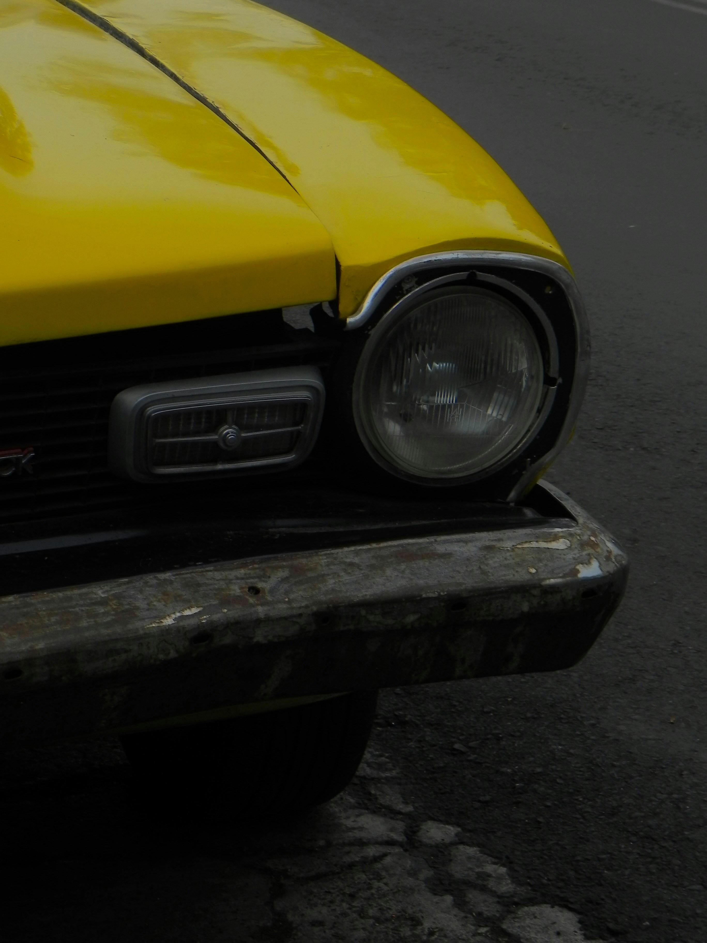 Close-up of the front end of a weathered yellow vintage car, emphasizing the headlight, grille, and worn bumper with a dark asphalt backdrop. A detailed automotive portrait showing aged chrome and paint wear.