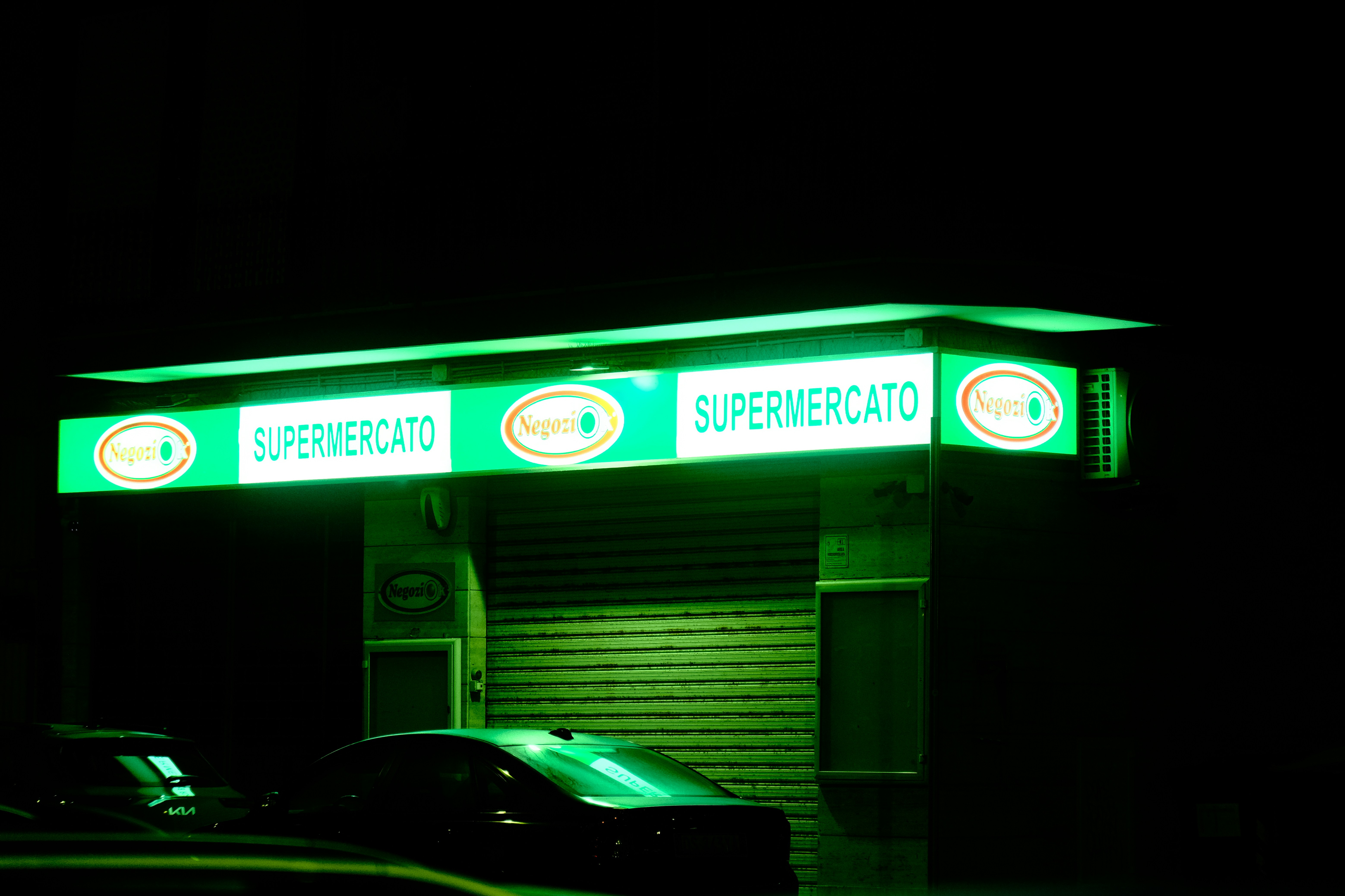 Neon-lit storefront sign of a quiet Italian supermarket glows green over a closed shutter, with two parked cars in silhouette beneath the sign.