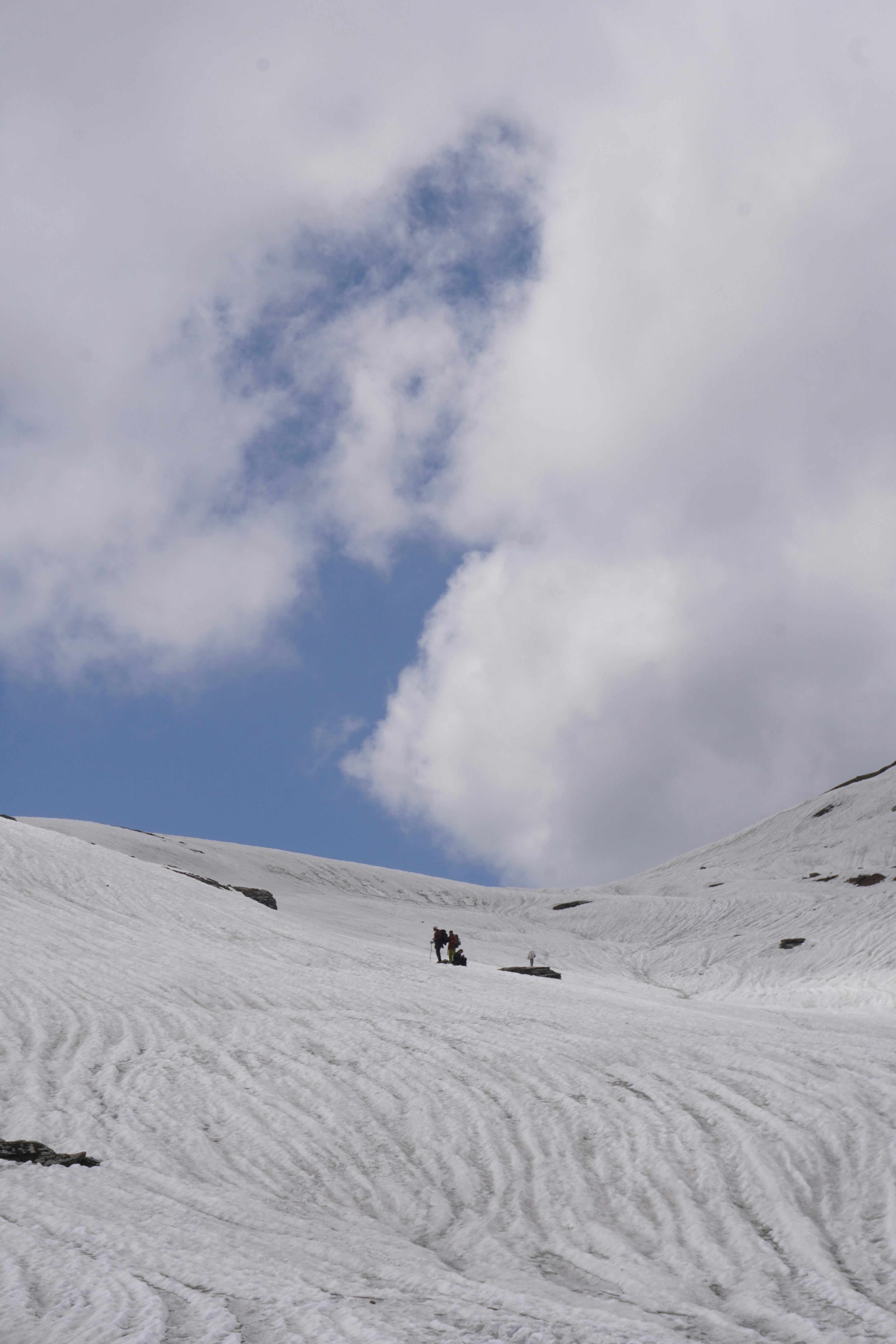 Snowy slope with two climbers near the center, small windbreak equipment visible; a patch of blue sky and clouds above, emphasizing scale and isolation.