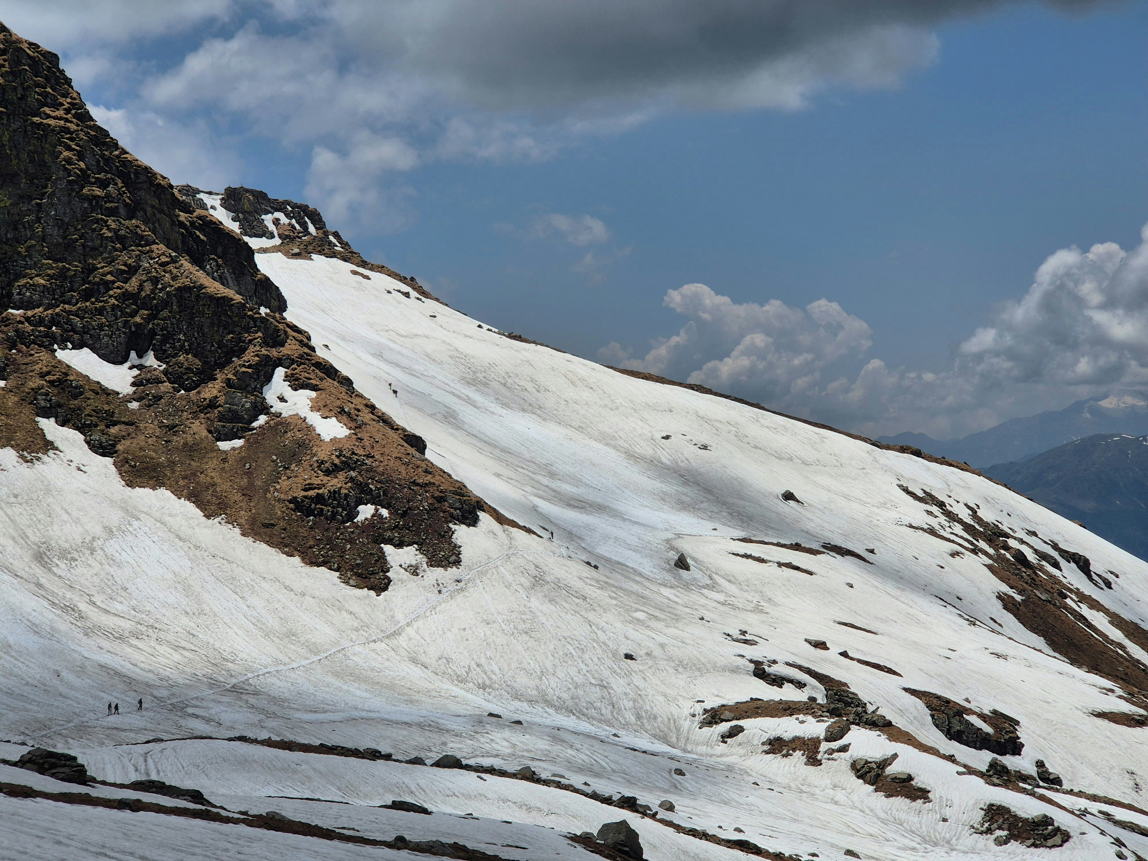 Snow-covered slope with rocky outcrops under a bright alpine sky, capturing the vast landscape and distant ridges.