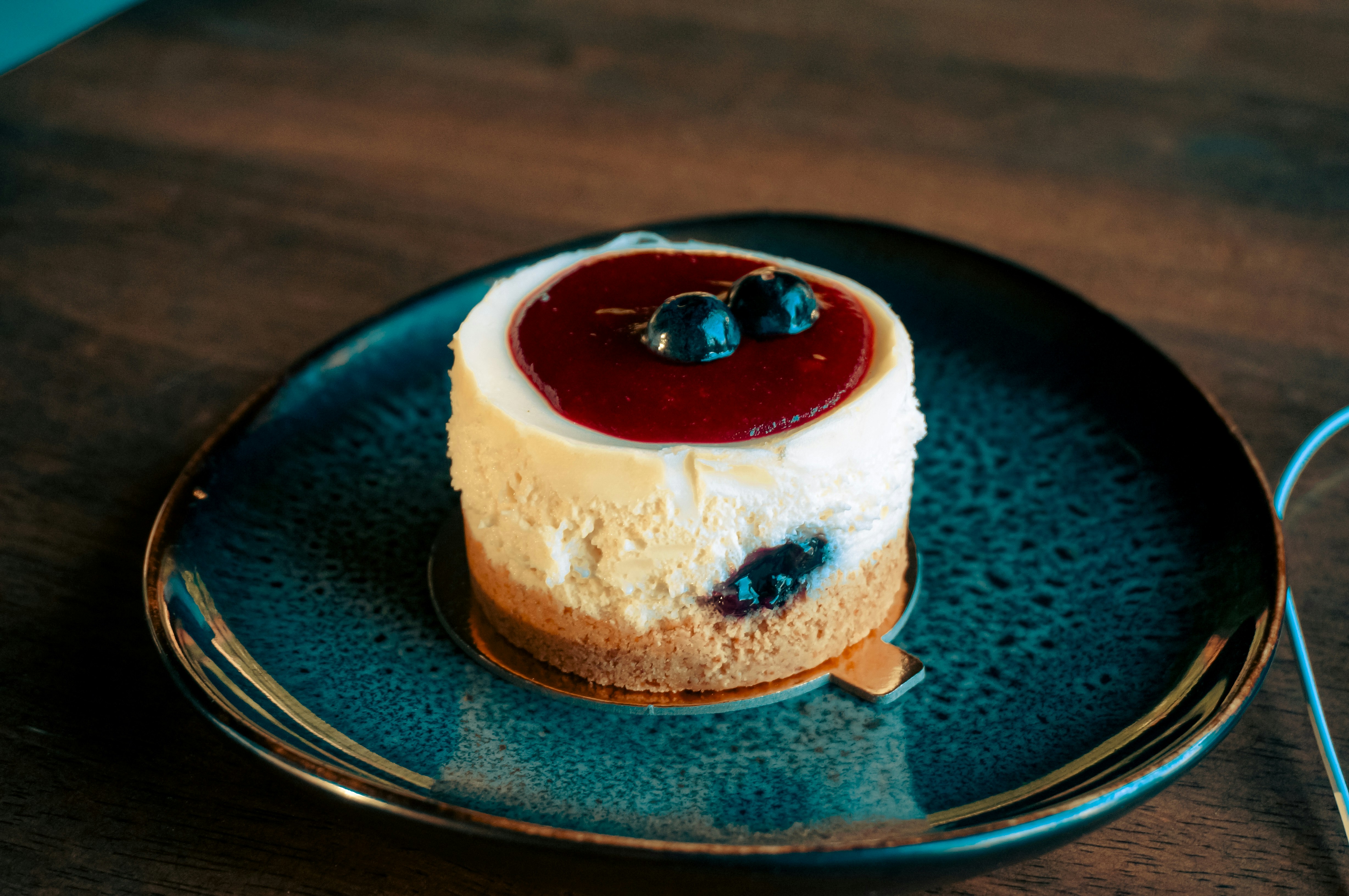 A small blueberry-topped cheesecake sits on a dark teal ceramic plate, plated on a wooden table. The dessert appears creamy with a berry glaze and a light crust, captured as a standalone confection.