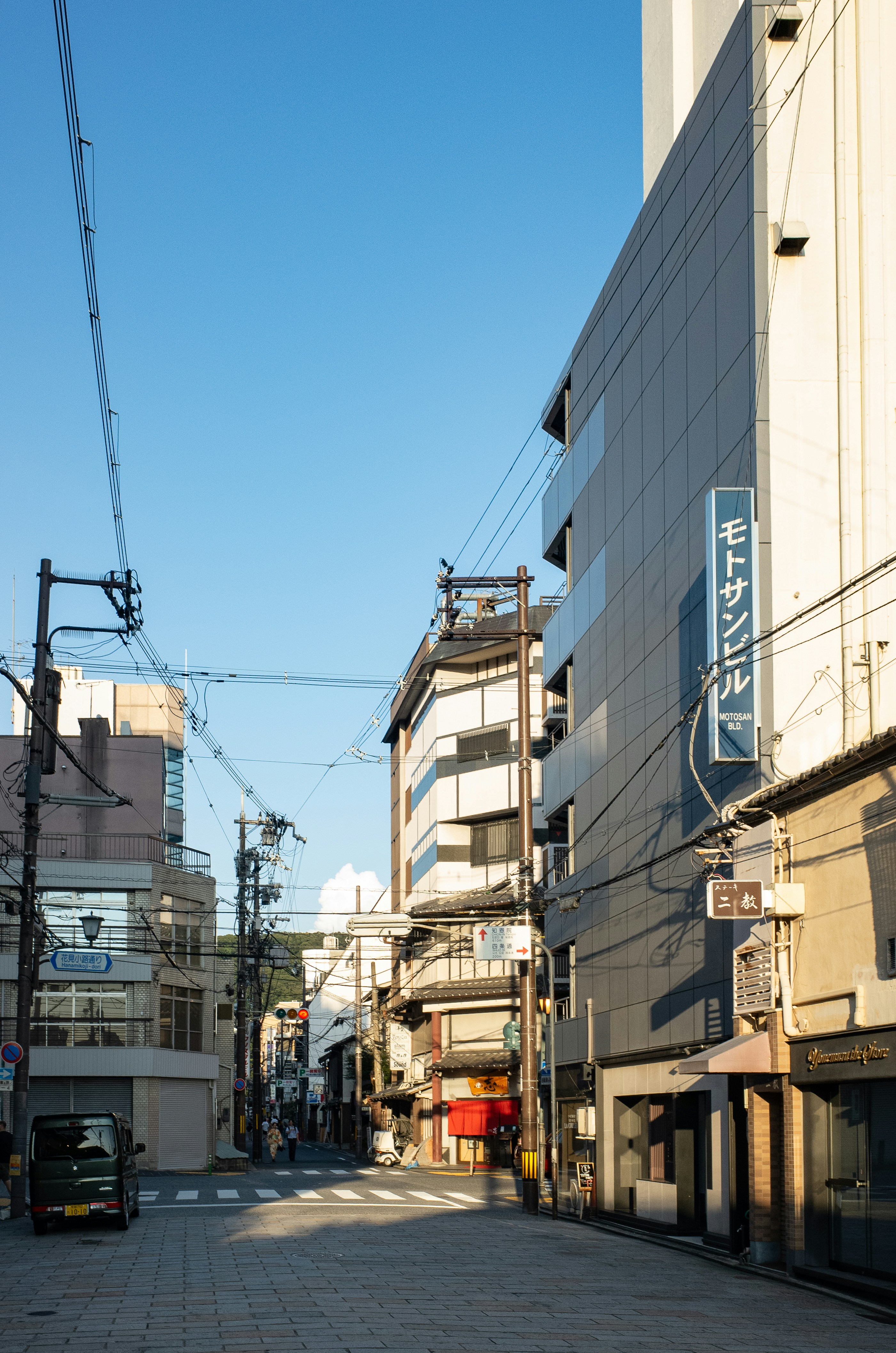 Sunlit city street with low-rise buildings and overhead power lines; a calm, empty pedestrian path leads toward a distant point of activity. Note: street scene photography.